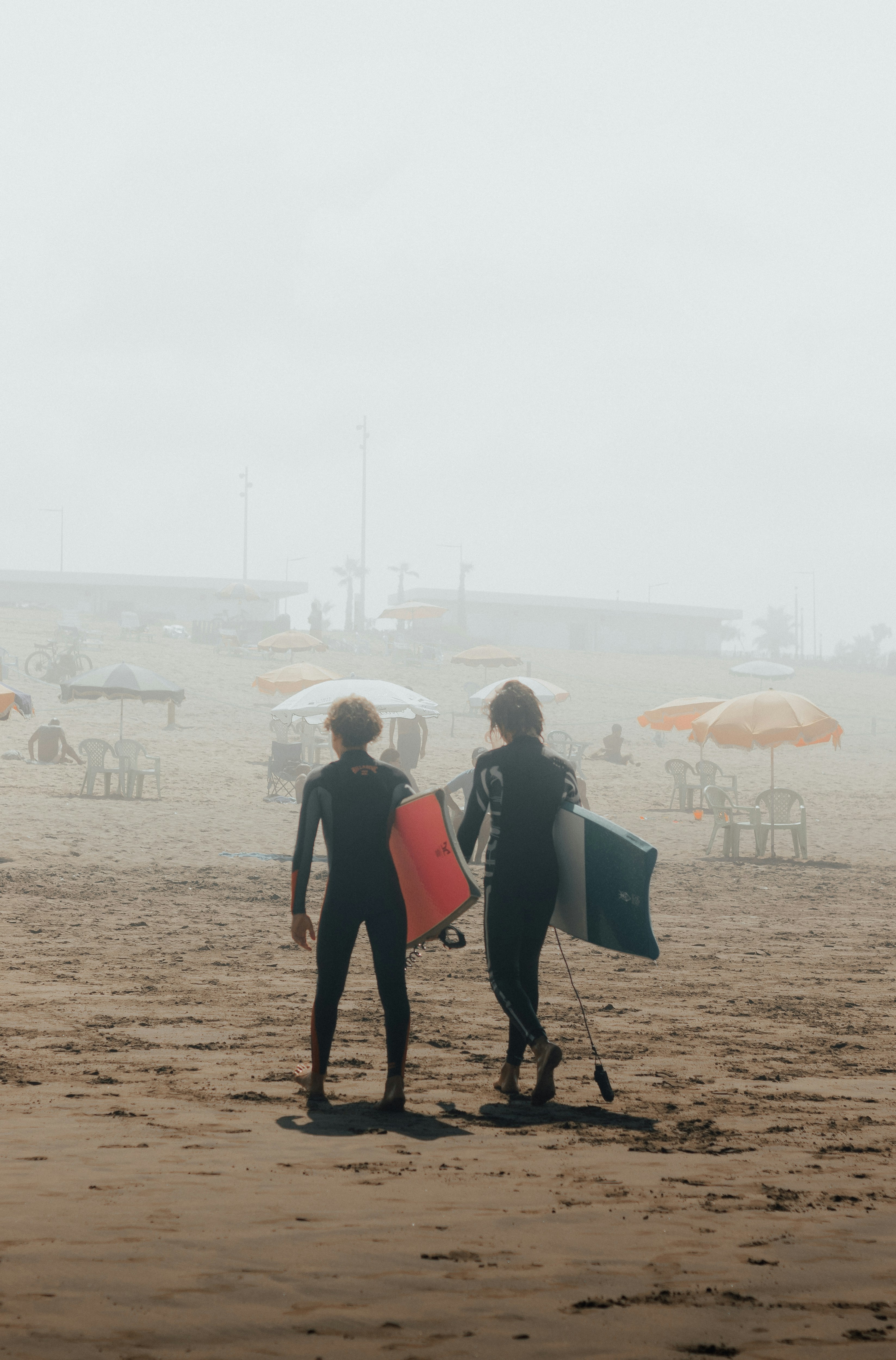 Two surfers in wetsuits walk towards the ocean, carrying their boards under a misty sky, with beach umbrellas dotting the sandy shore.