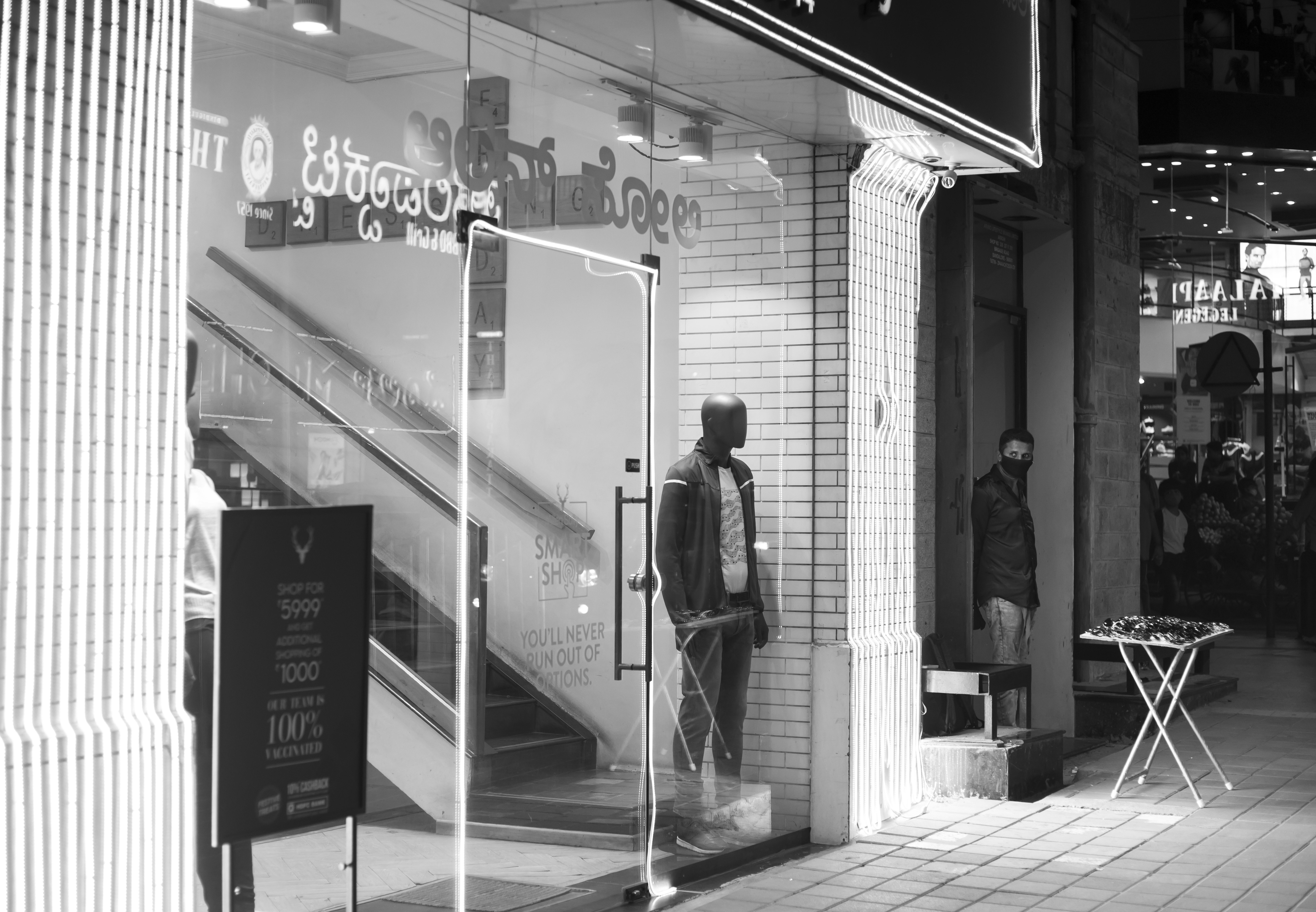 A mannequin dressed in casual attire stands in a storefront, while a person observes nearby, creating a juxtaposition of stillness and life.