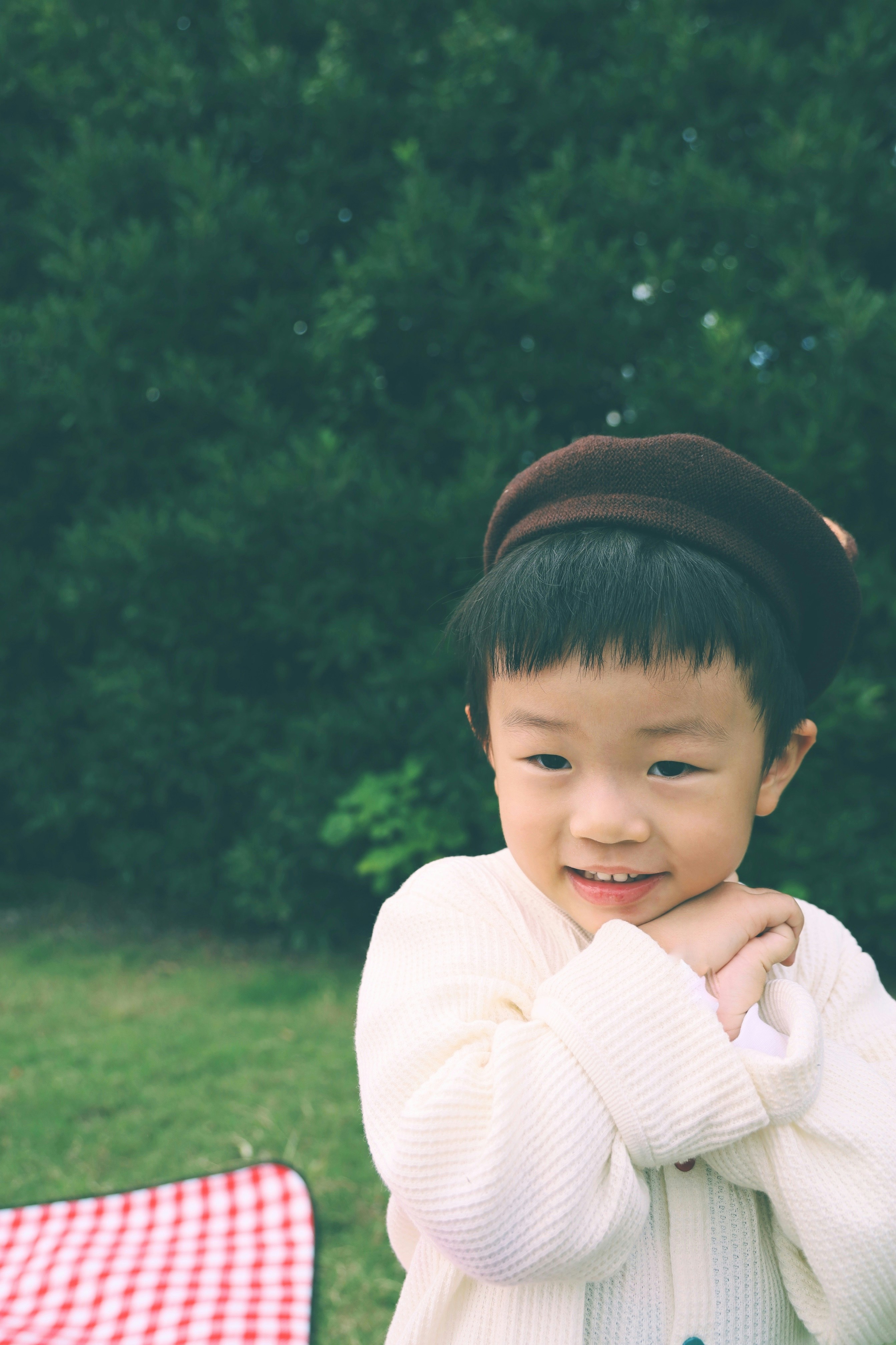 Young child in a cozy sweater and hat smiles playfully in a green outdoor setting, evoking a sense of innocence and joy.