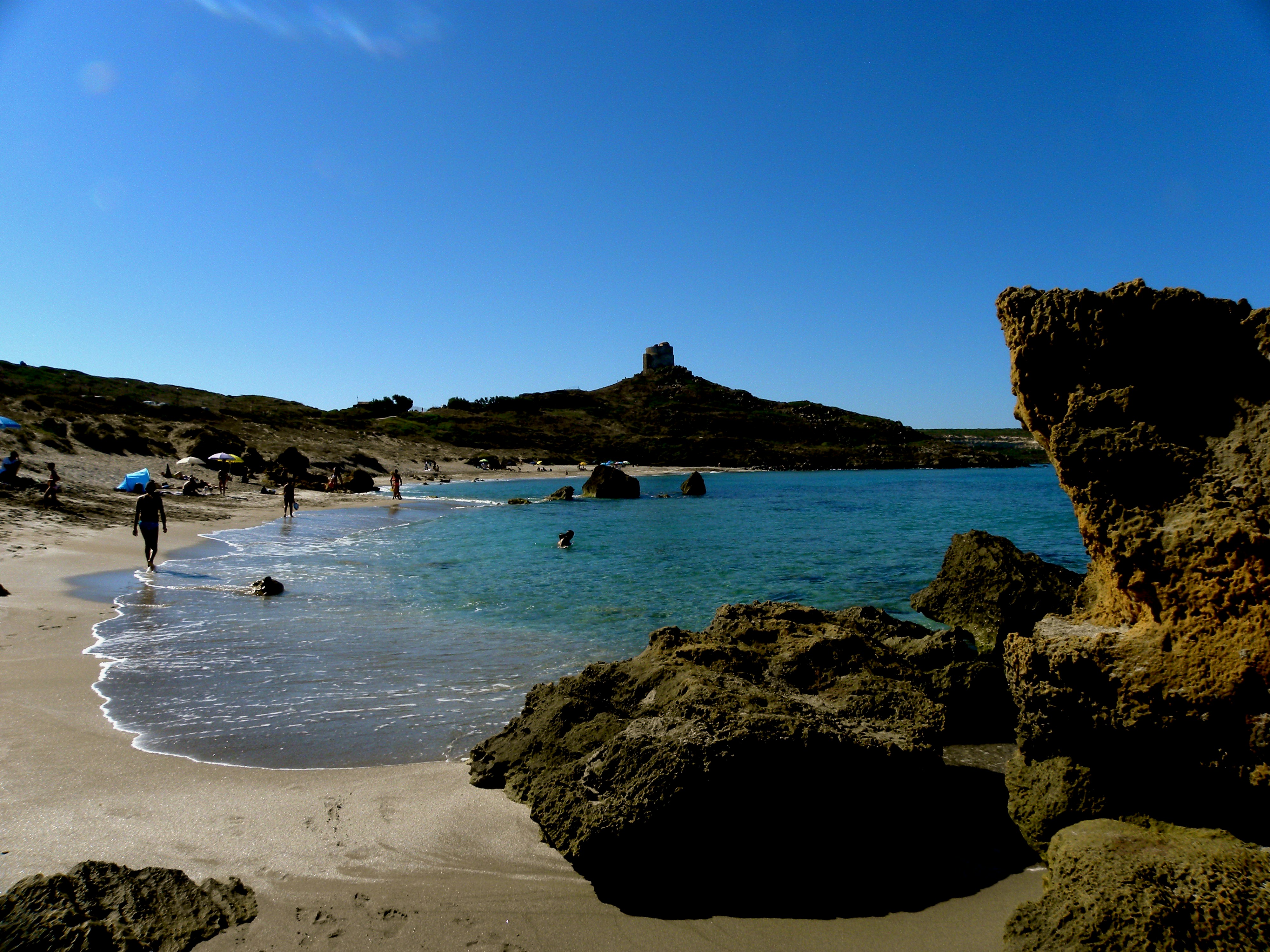 Beachgoers enjoying a sunny day by the rocky coastline with clear blue skies.