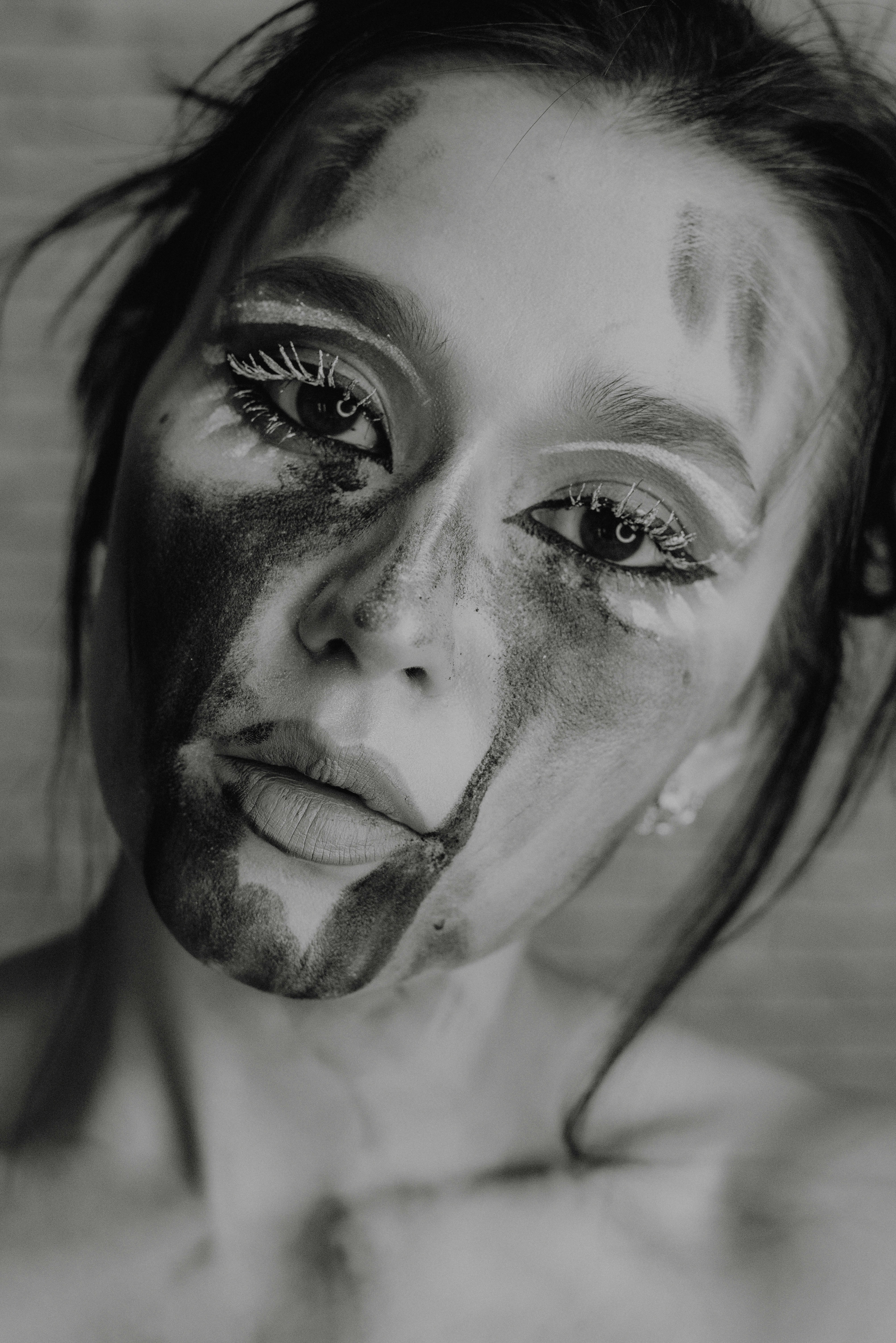 Artistic portrait featuring a model with striking facial paint and dramatic makeup, emphasizing emotional depth and contrast. Black and white tones enhance the visual impact.
