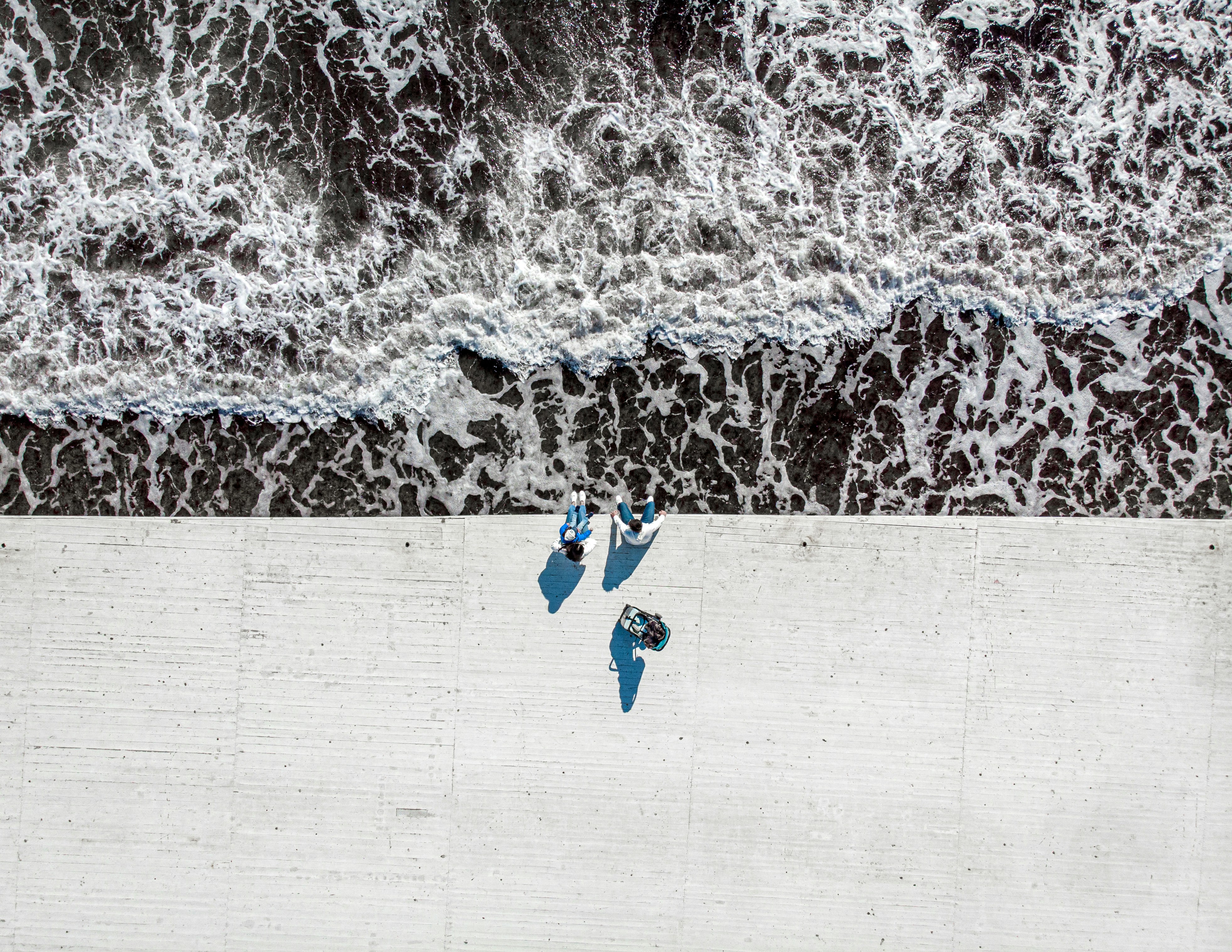 Three figures stand on a concrete platform as waves crash nearby, creating a dynamic contrast between land and sea.