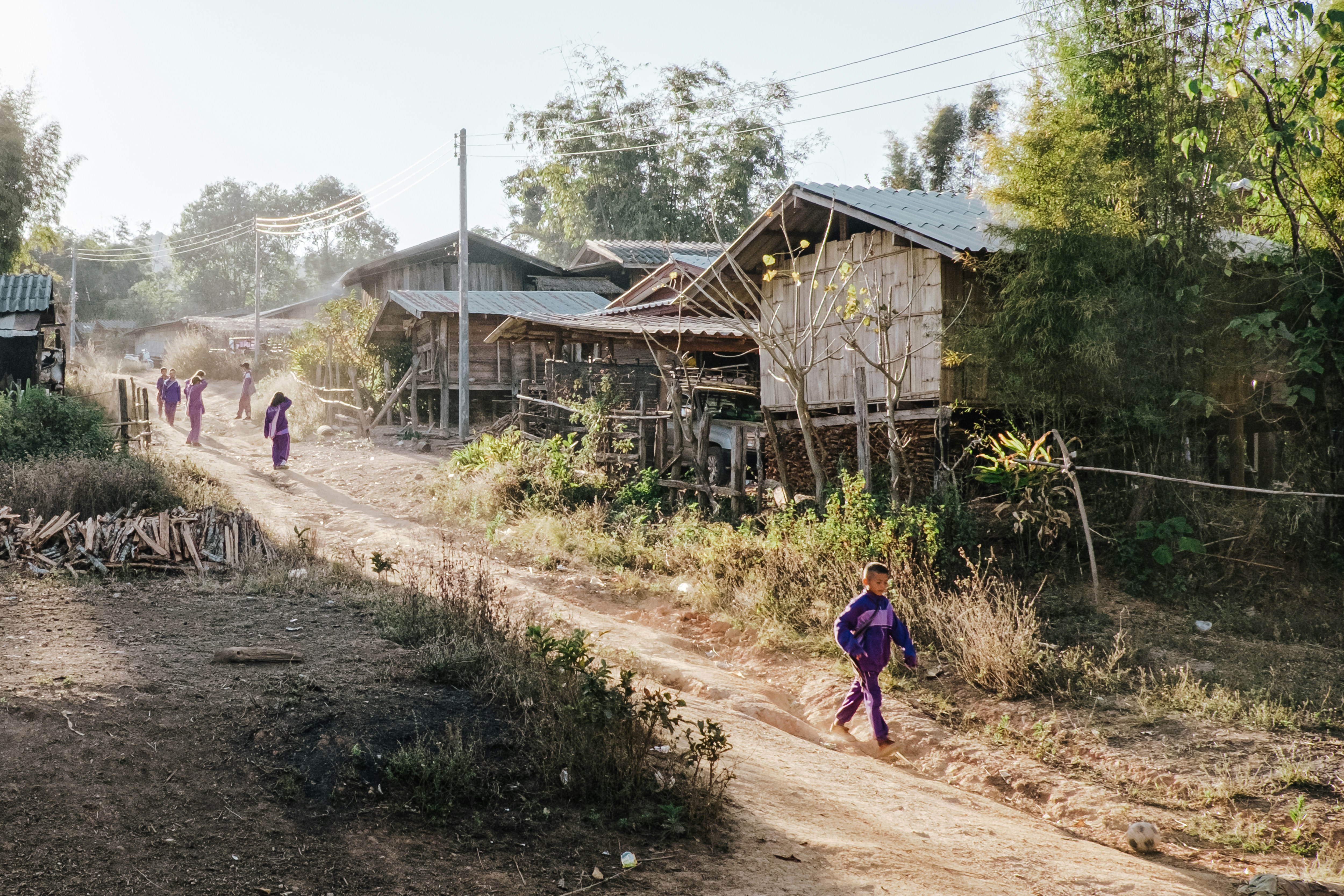 Children in purple uniforms walk along a dirt path lined with rustic houses and greenery in a rural Thai village.