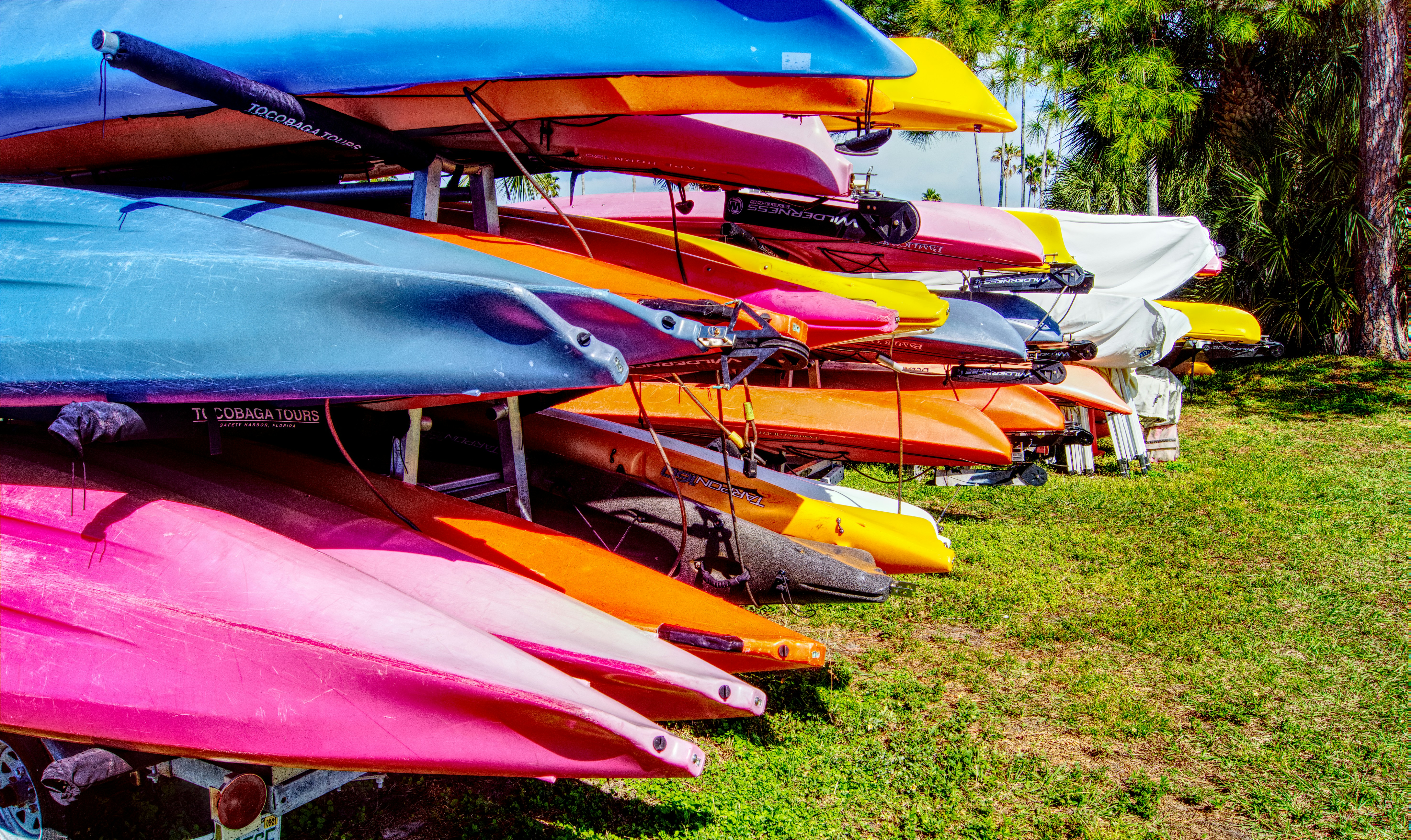 Rows of colorful kayaks stacked neatly on green grass under bright sunlight.