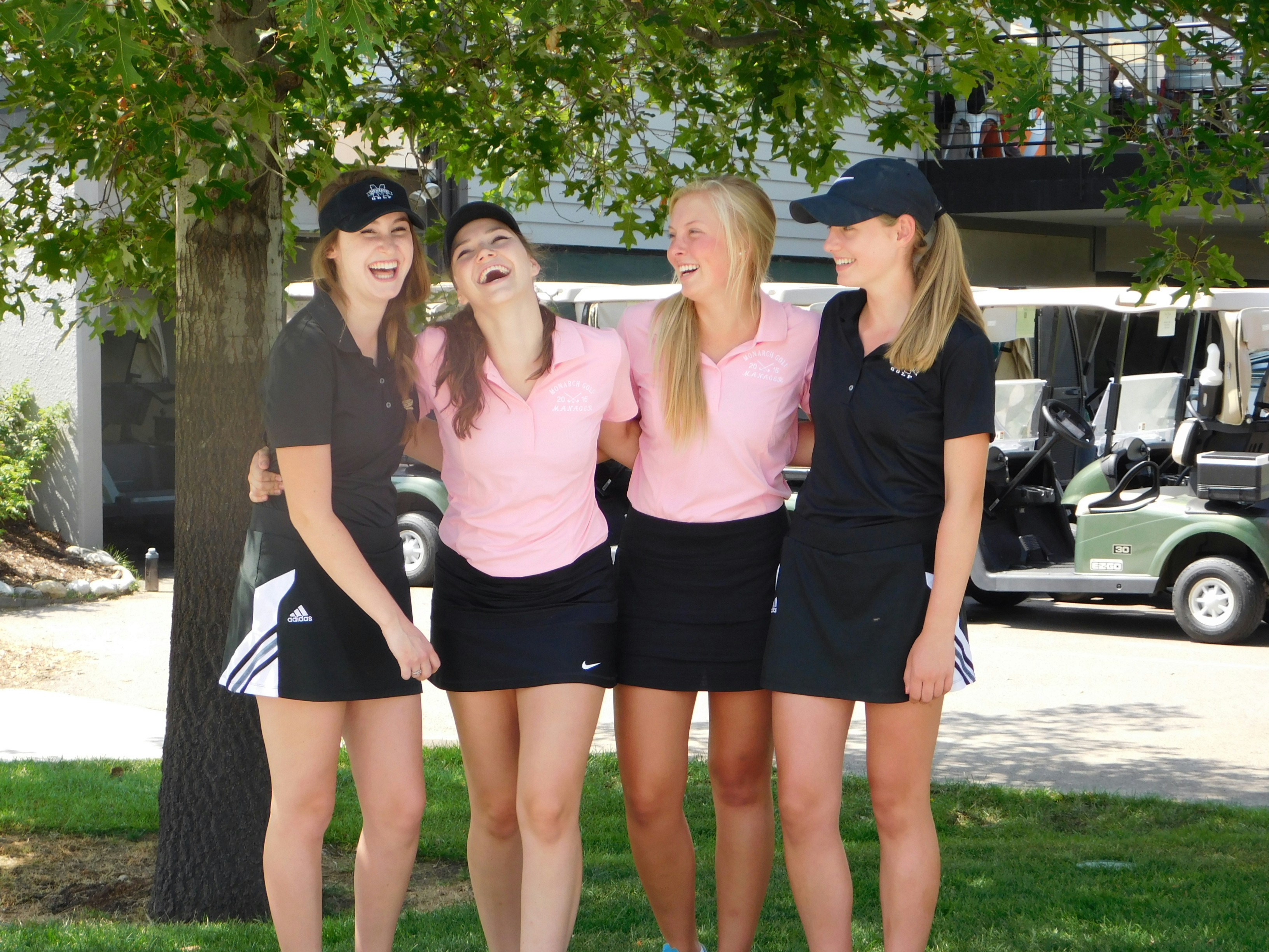 Group of four young women in golf attire sharing a laugh under a tree with golf carts in the background.