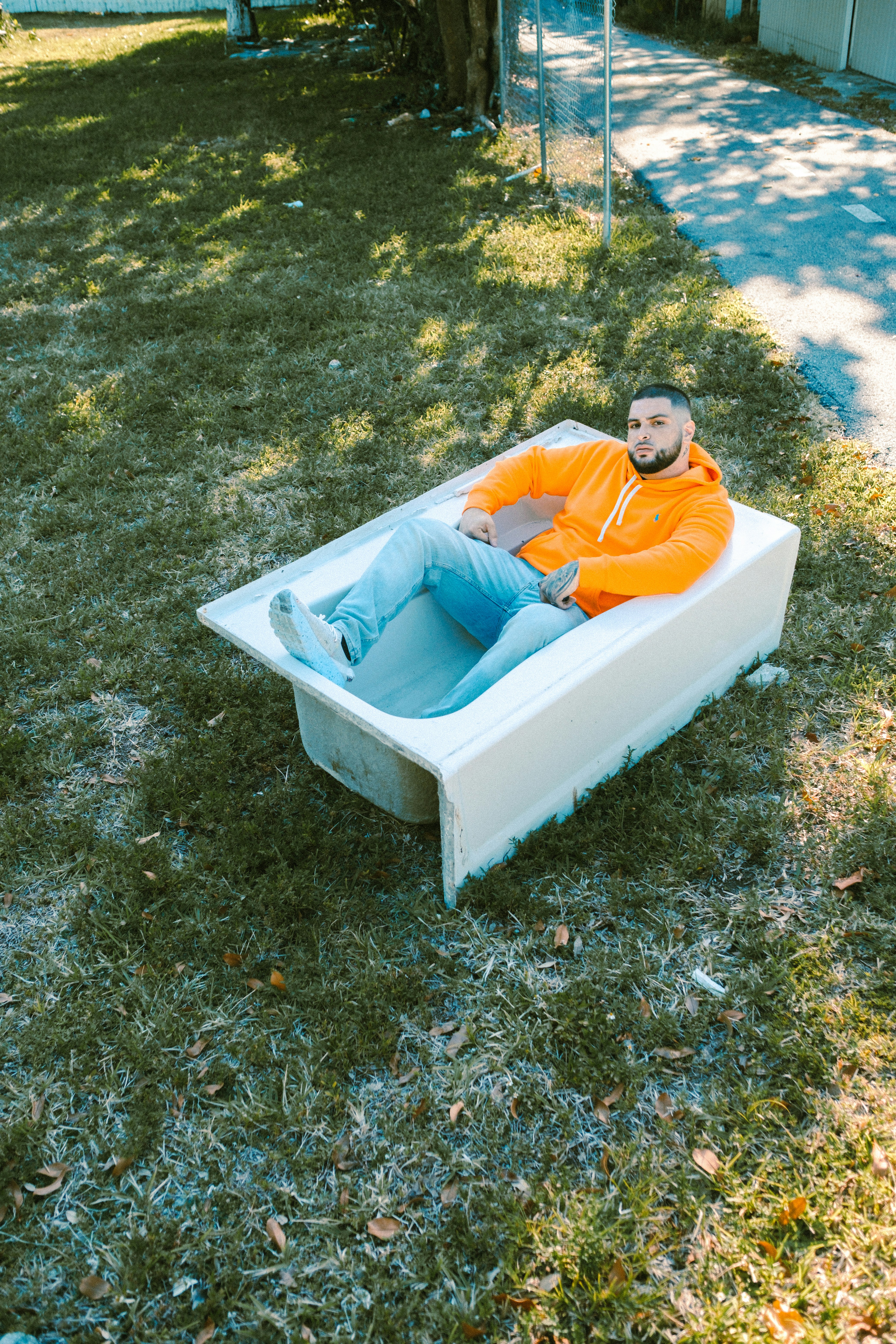 Man in Bright Orange Hoodie Relaxing in a White Bathtub Set in a Sunlit Yard