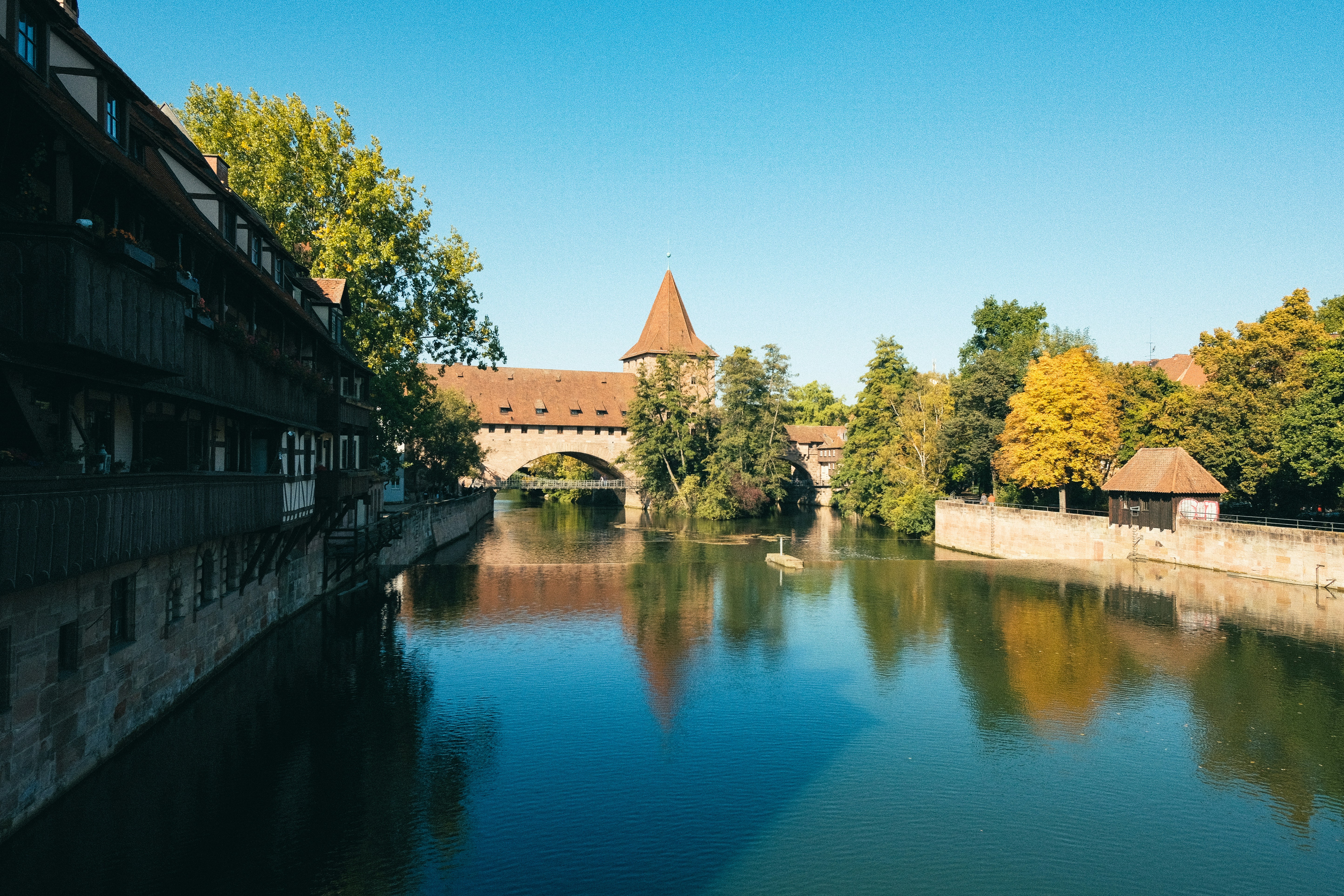 Stone bridge over a tranquil river with lush trees and historic architecture under a clear blue sky.