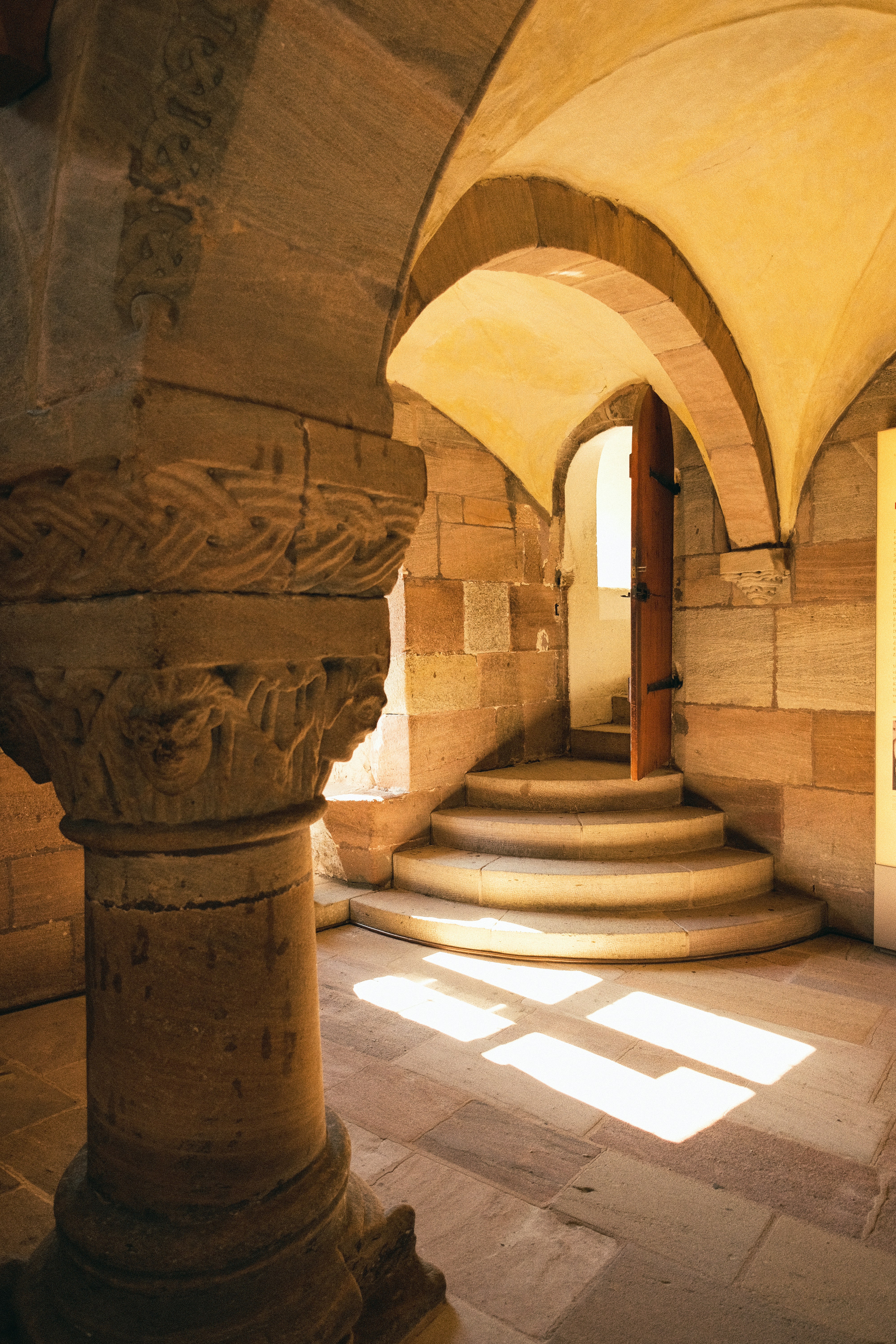 Intricate stone column leads to a softly lit staircase and an inviting wooden door, revealing a glimpse of architectural elegance within a historic space.