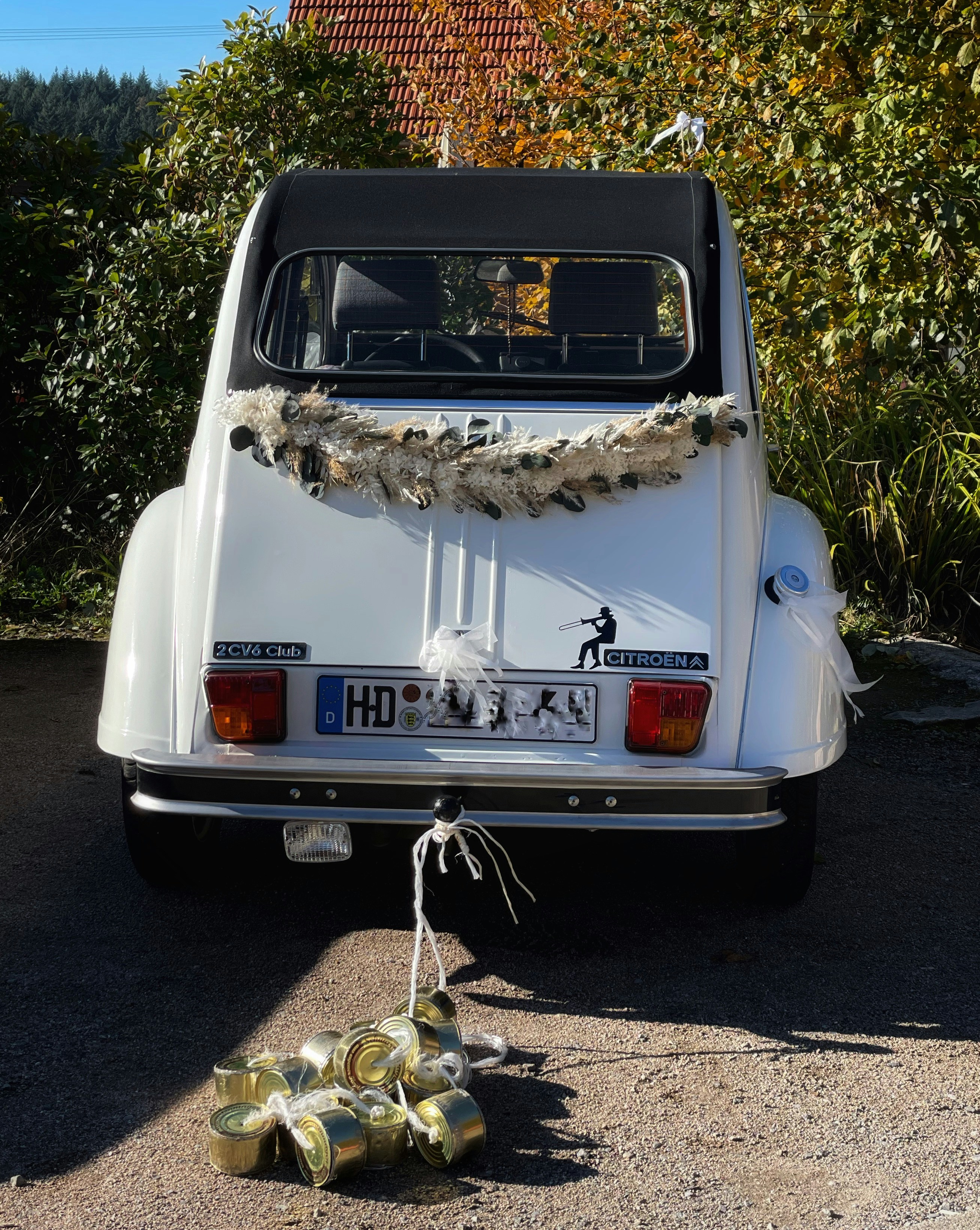 Rear view of a decorated Citroën 2CV, featuring a garland and trailing cans, set against a backdrop of vibrant autumn foliage.