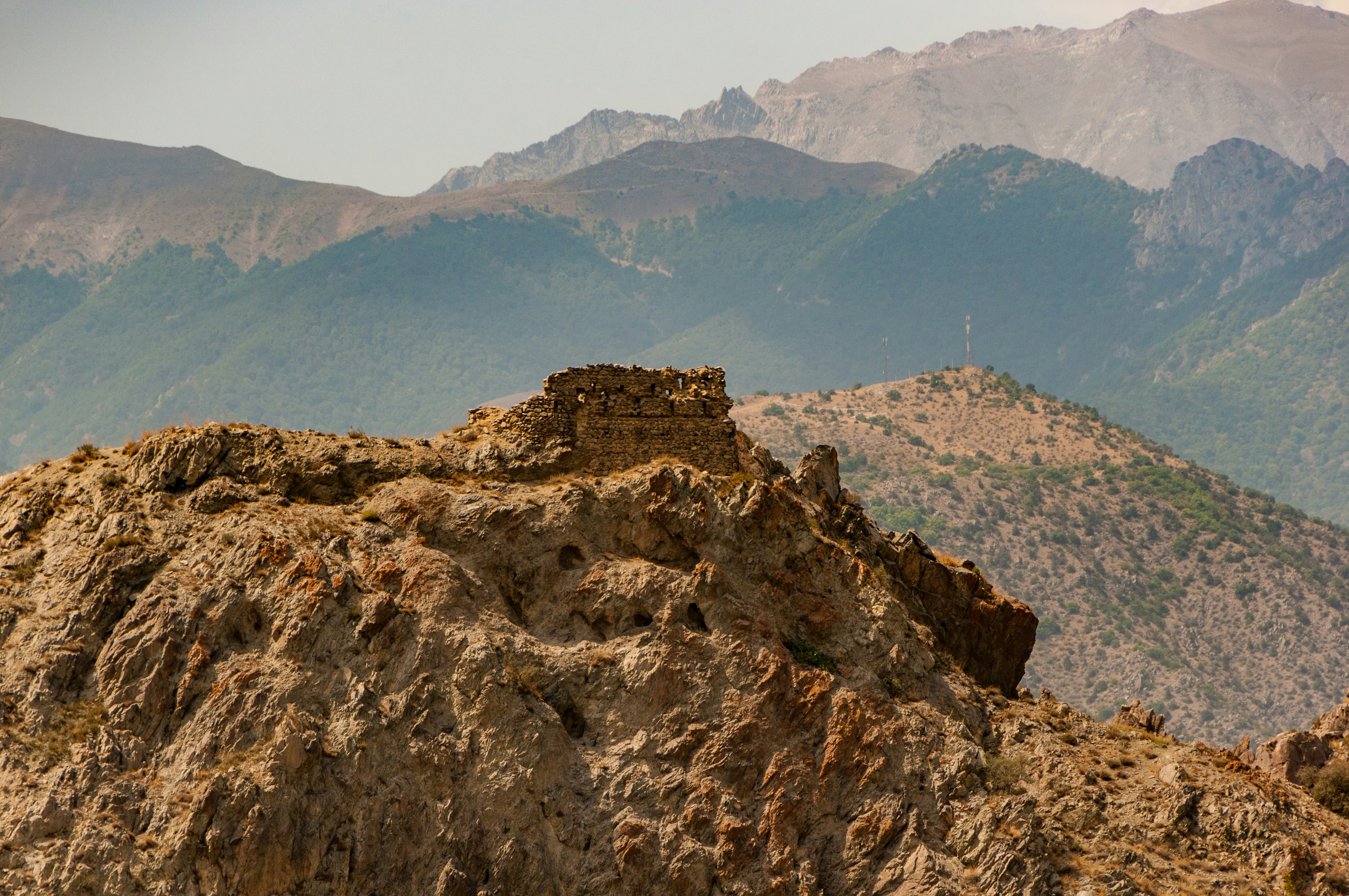 Weathered stone ruins perched atop a rugged mountain, surrounded by a vast landscape of rolling hills and distant peaks.