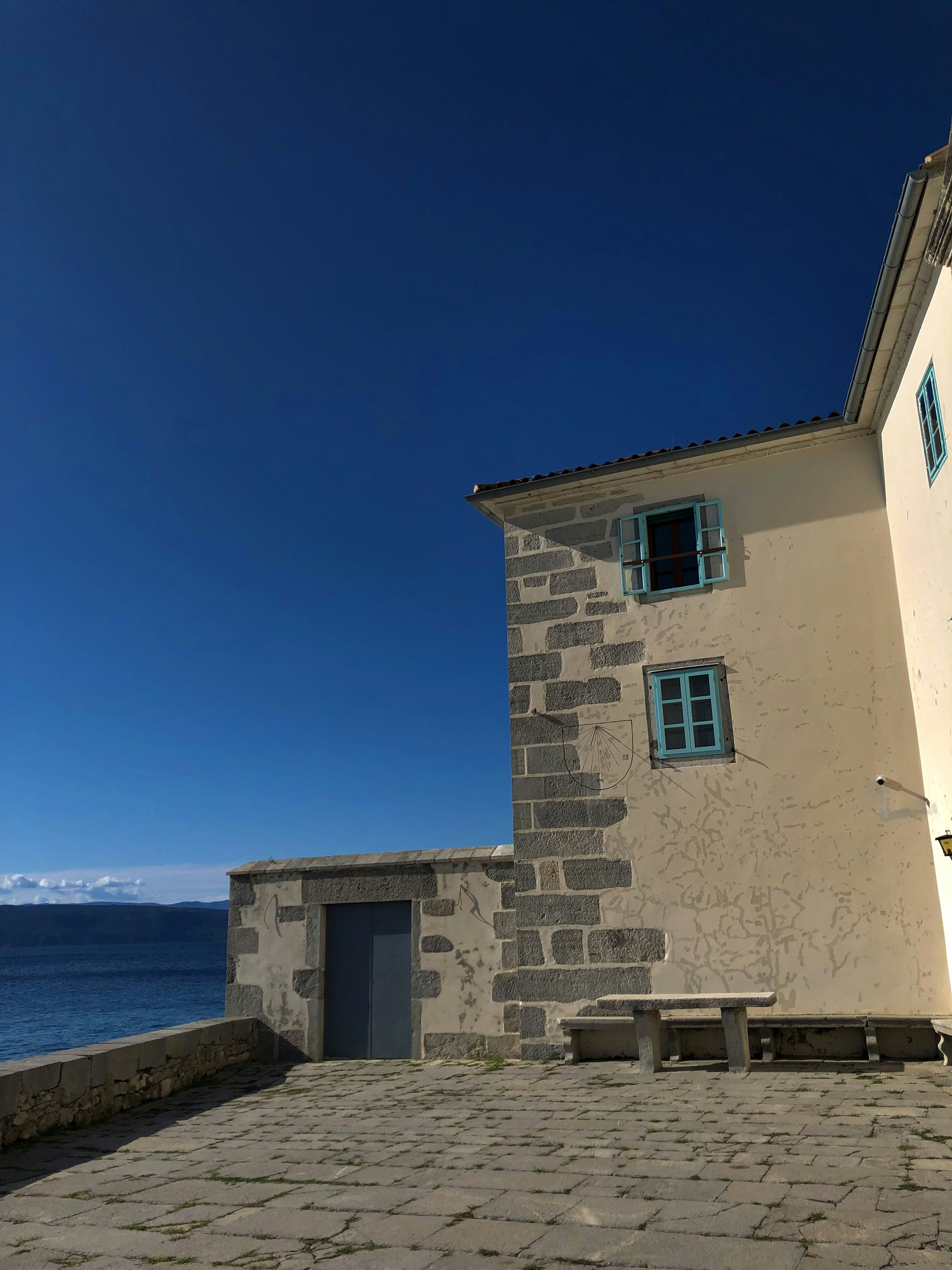 Charming coastal building with turquoise windows under a clear blue sky, overlooking the sea. The structure's weathered texture contrasts beautifully with the vibrant surroundings.
