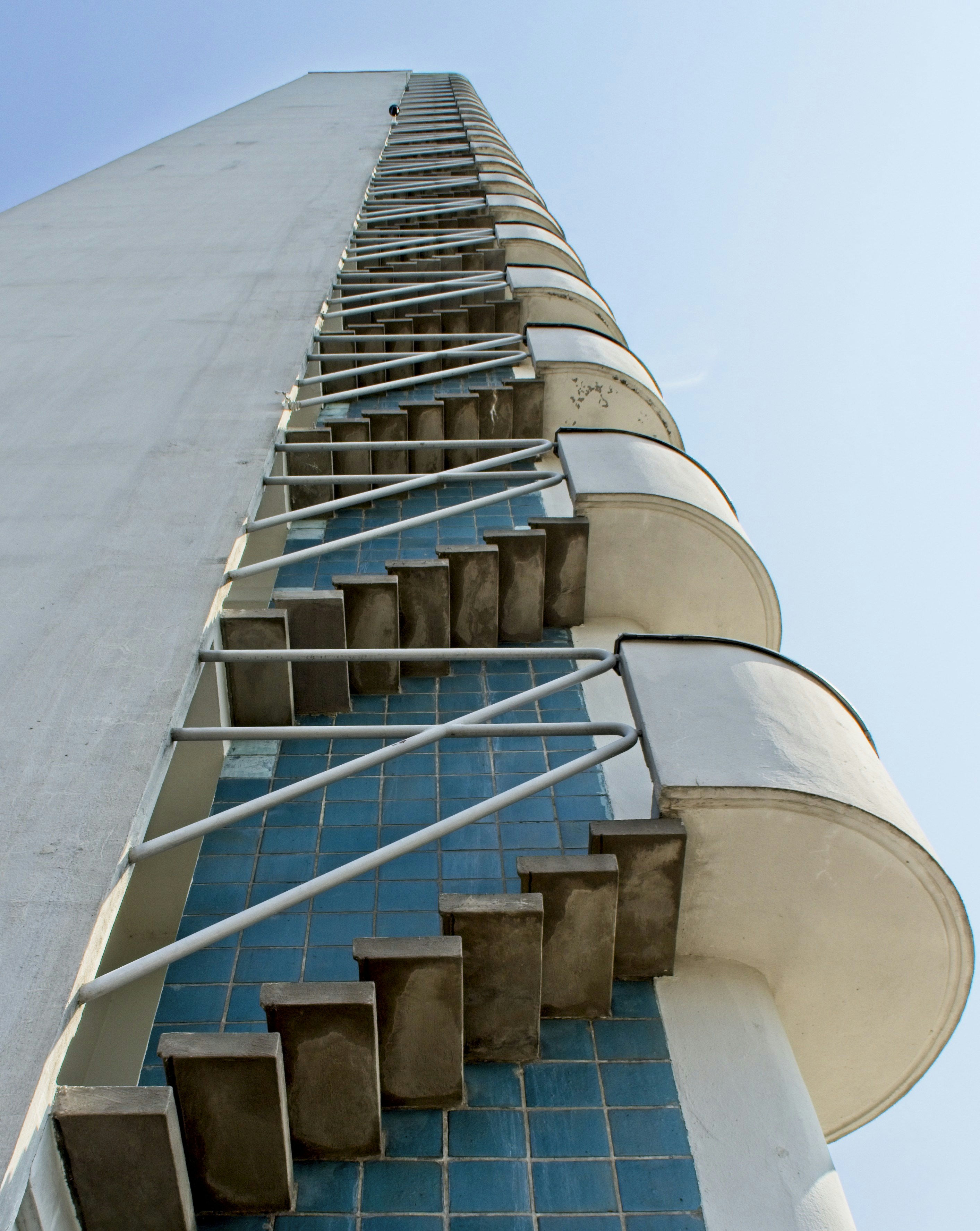 Vertical concrete tower with external curved staircases climbing along its side against a clear blue sky. The composition emphasizes geometric repetition and architectural detail.