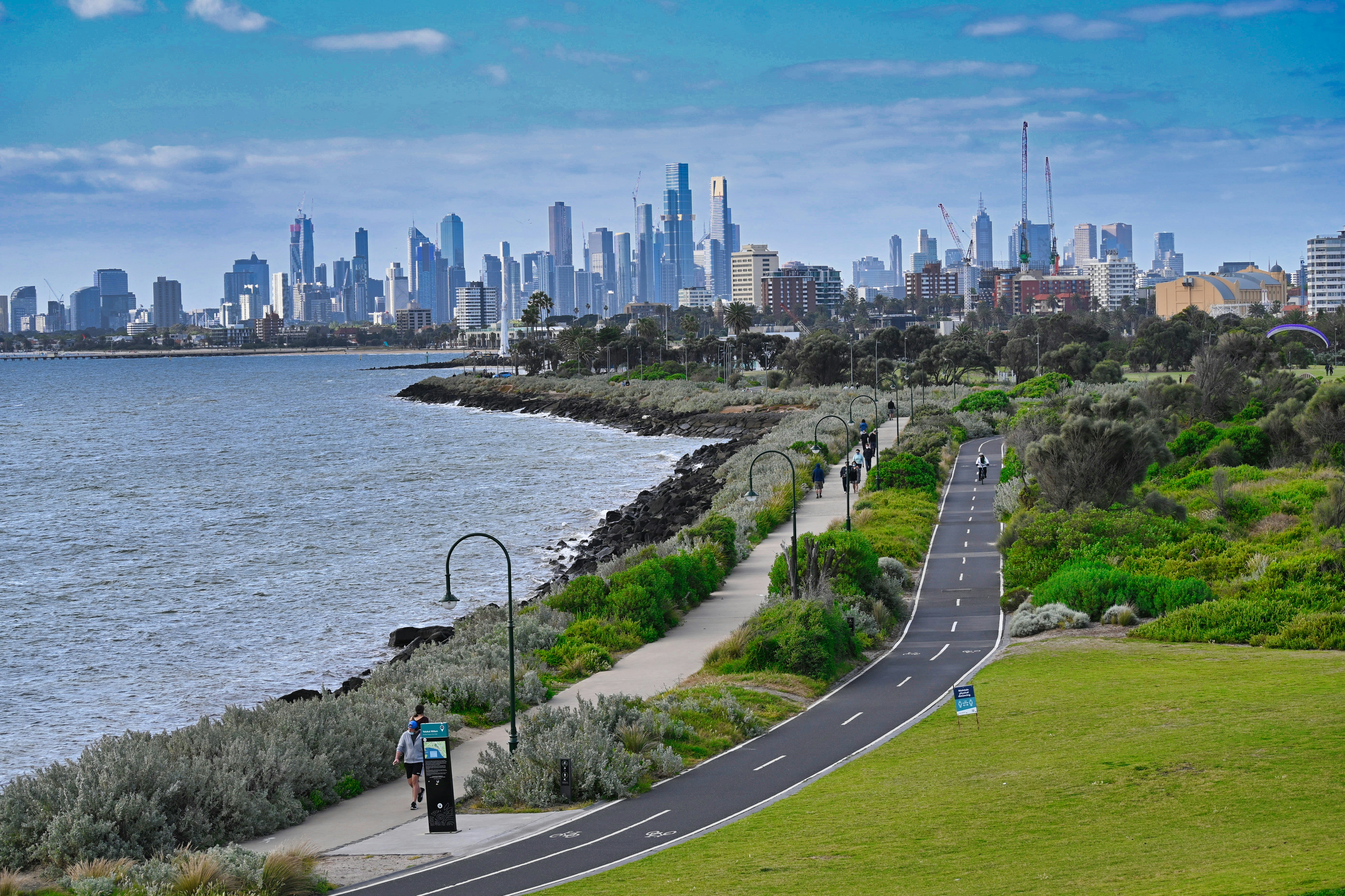 Coastal pathway lined with greenery leading towards a vibrant city skyline under a clear blue sky.