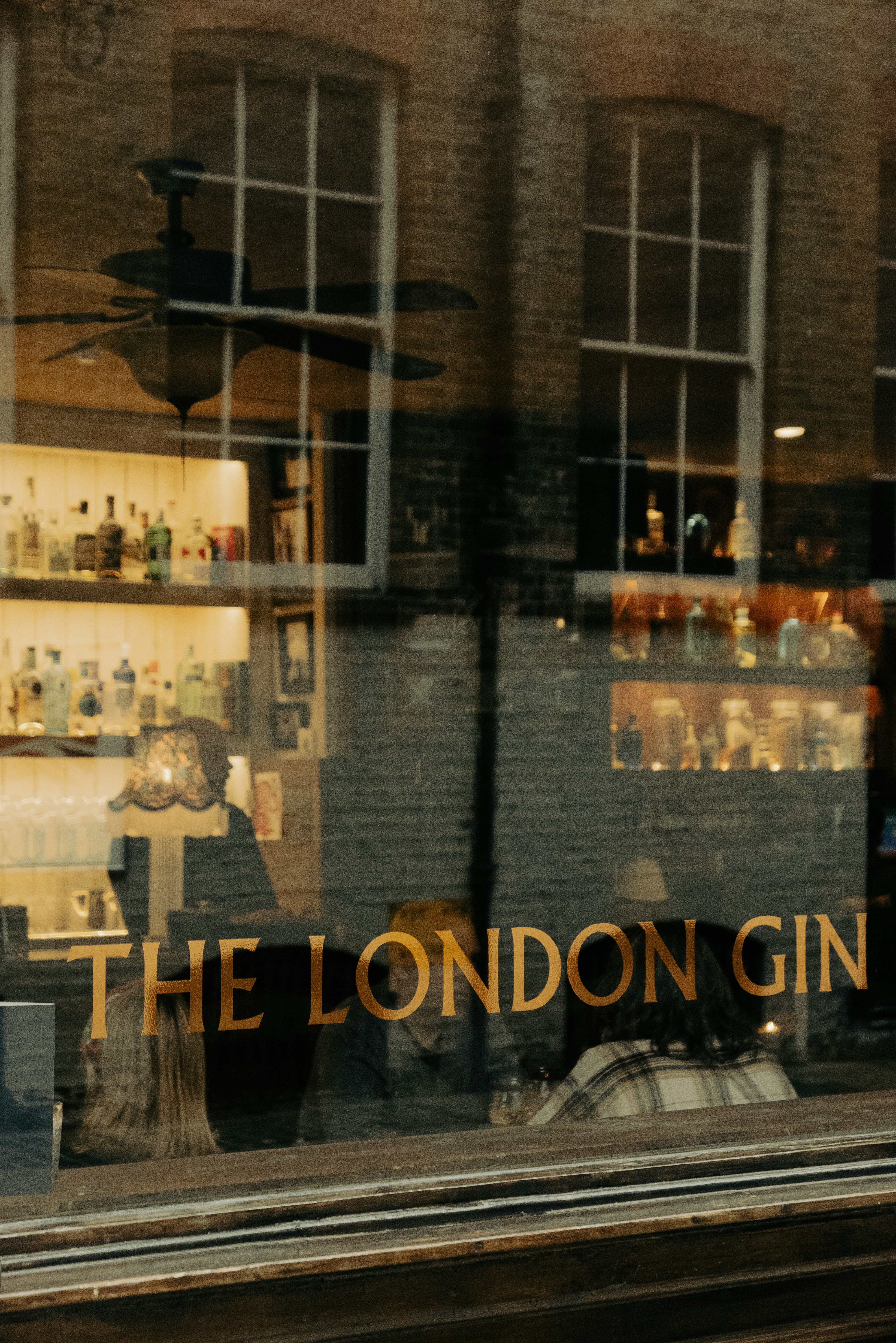 Window reflection shows a London gin bar with backlit bottles on shelves and warm interior lighting, with the 'THE LONDON GIN' signage across the lower pane.