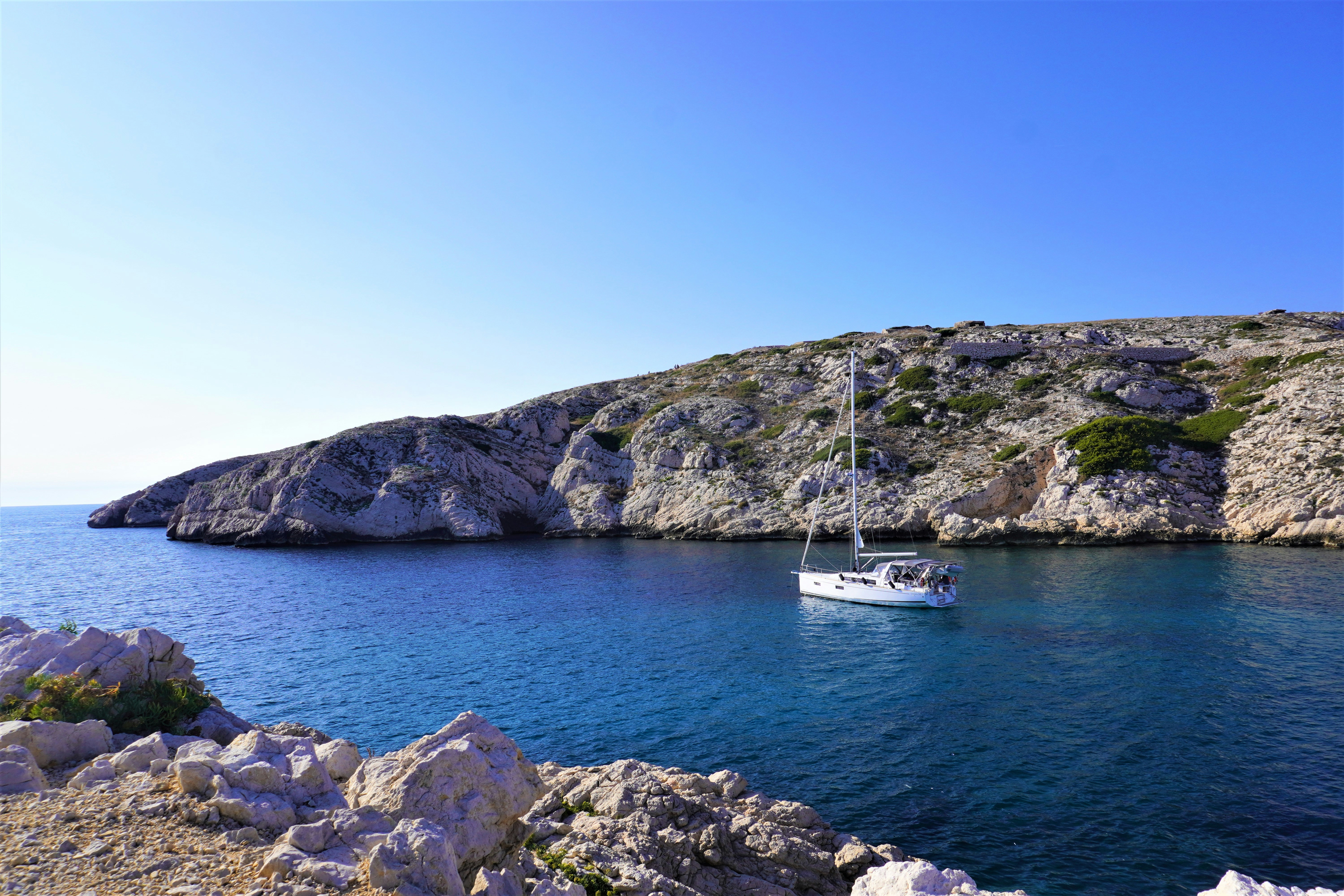 White sailboat anchored in a tranquil blue cove surrounded by rugged cliffs under a clear sky.