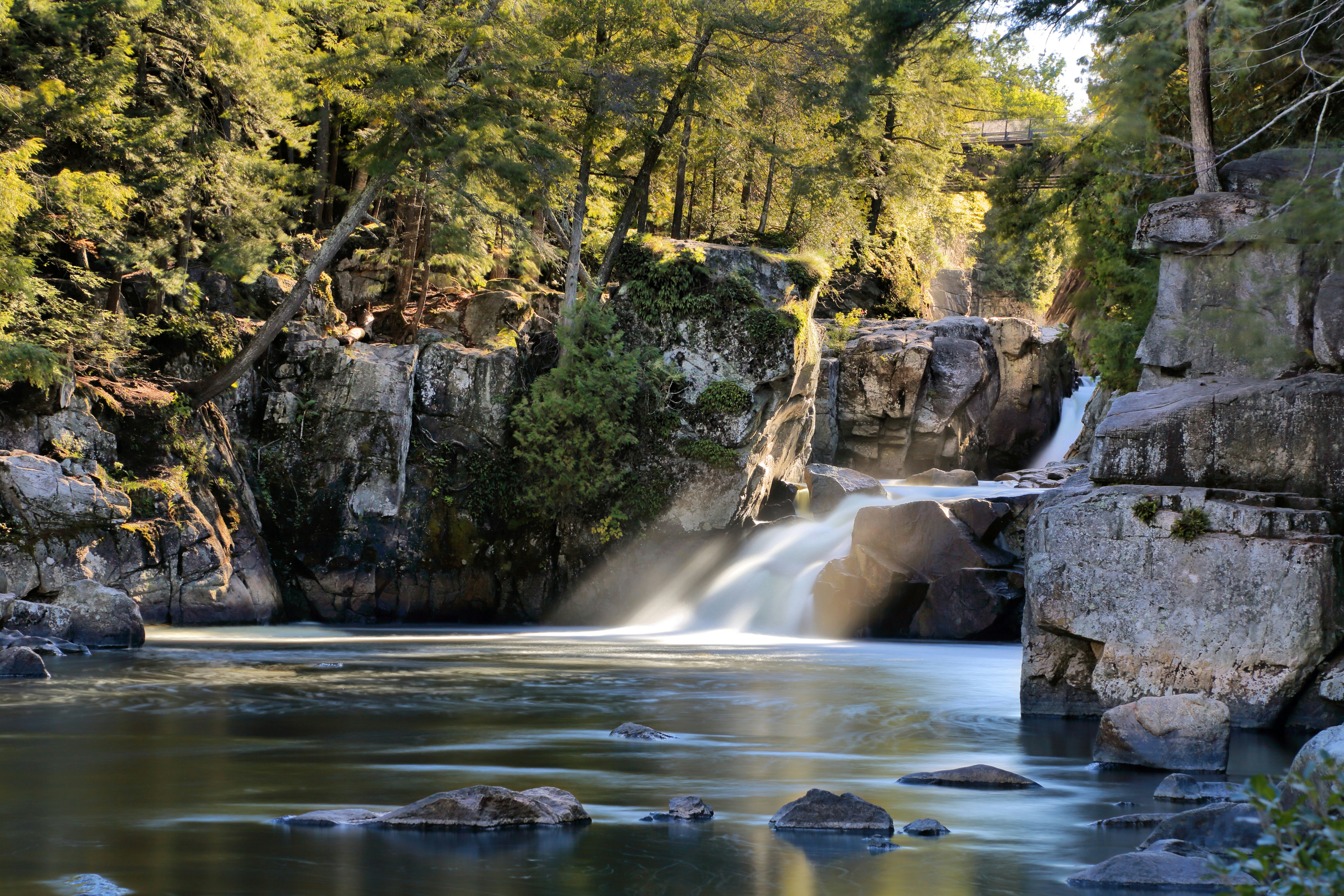Gentle waterfall flowing through rocky terrain, surrounded by lush greenery and dappled sunlight. The serene water reflects the tranquility of the natural landscape.