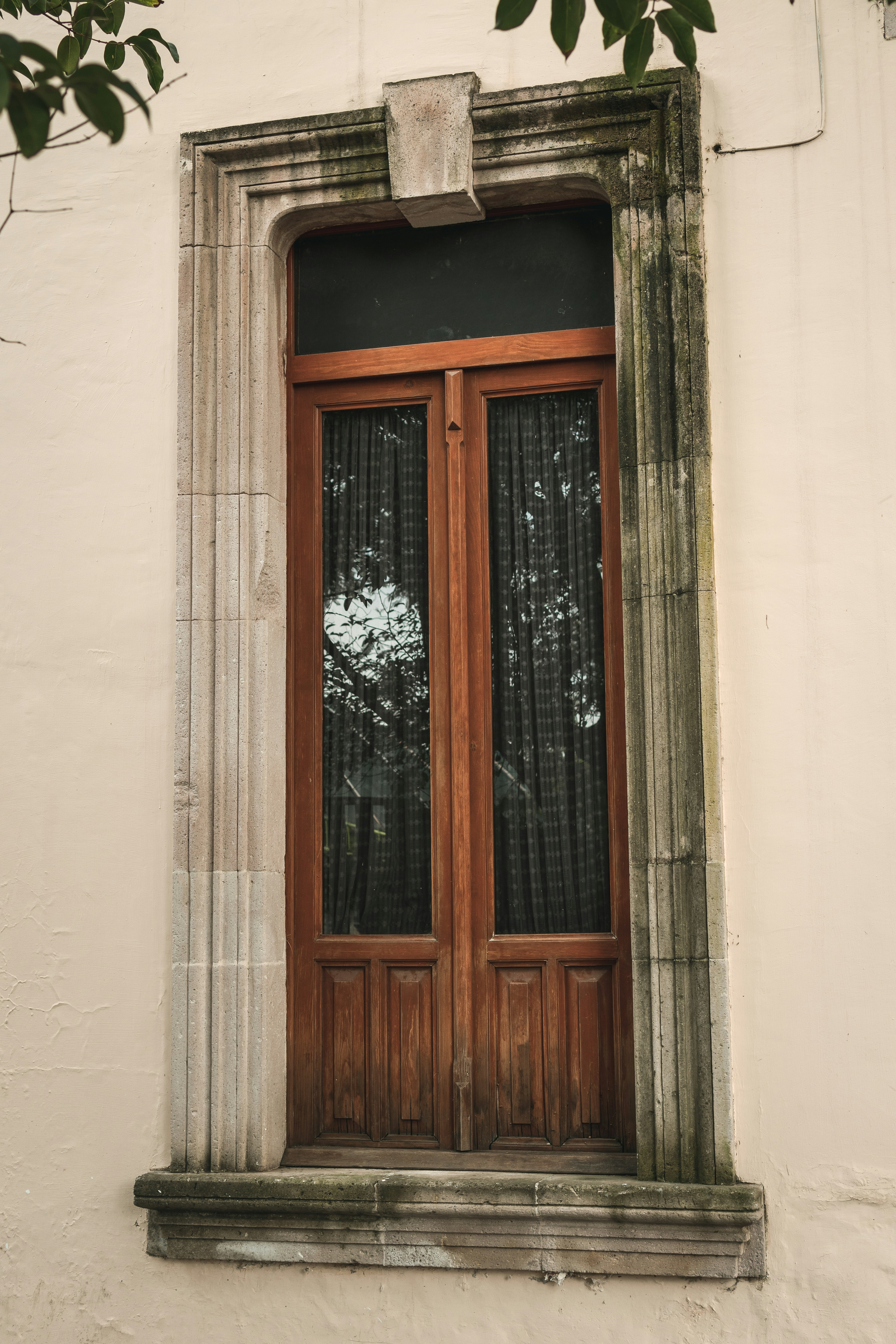 Elegant wooden window framed by intricate stonework, showcasing patterned curtains behind glass. A glimpse of nature is reflected in the window.