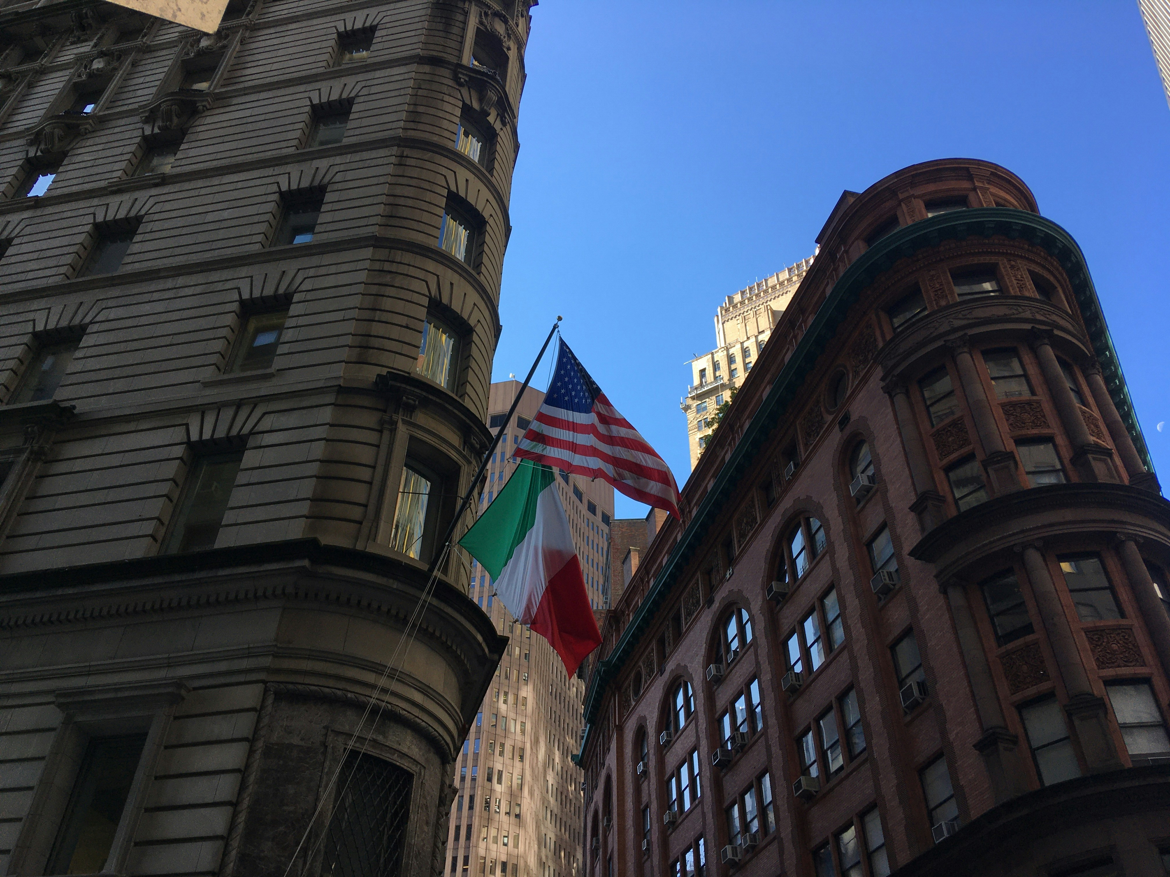 Flags of the United States and Italy fluttering between historic buildings in an urban setting.