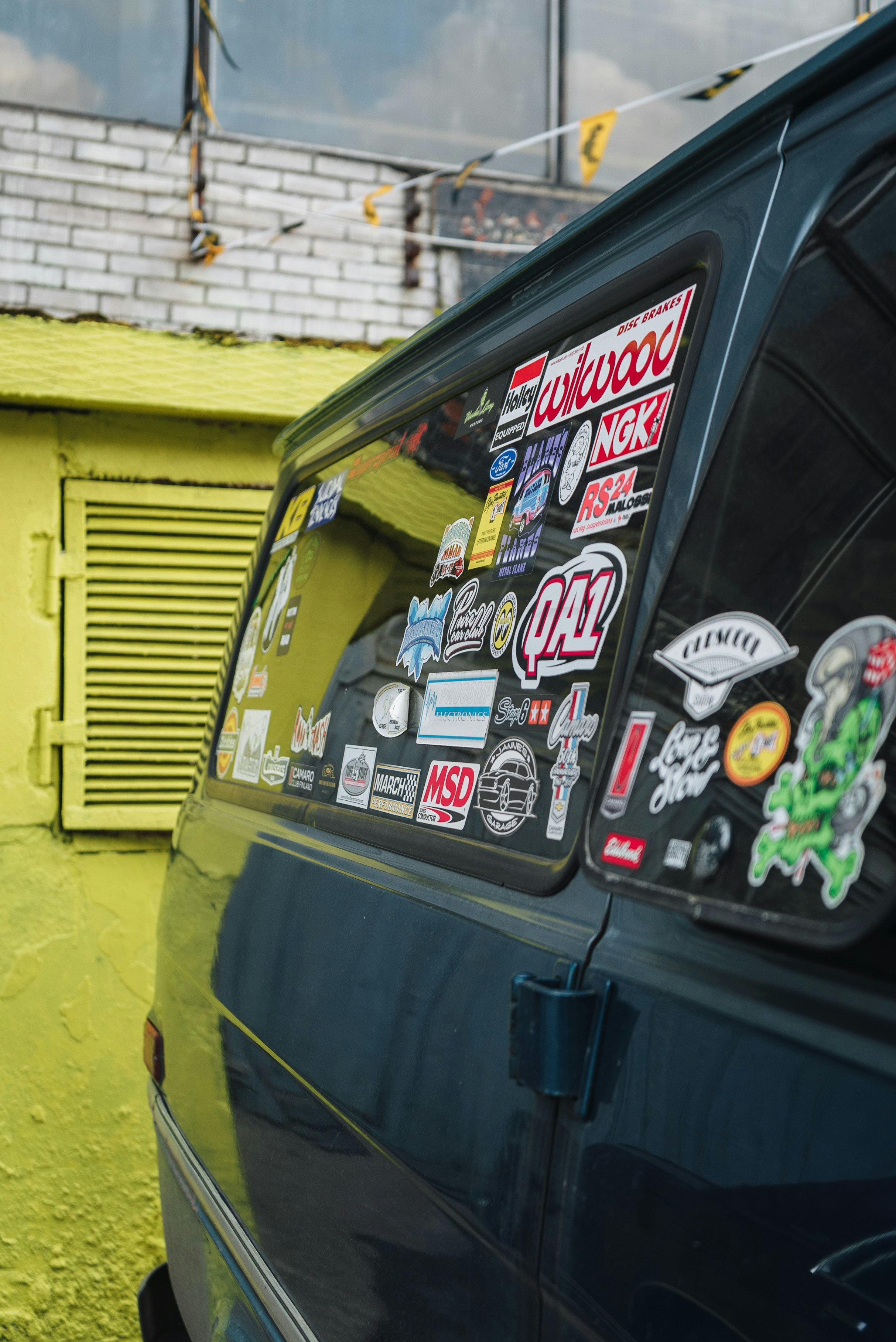 Colorful stickers adorn the side of a vintage van, set against a vibrant yellow wall. The scene captures a blend of nostalgia and urban art.