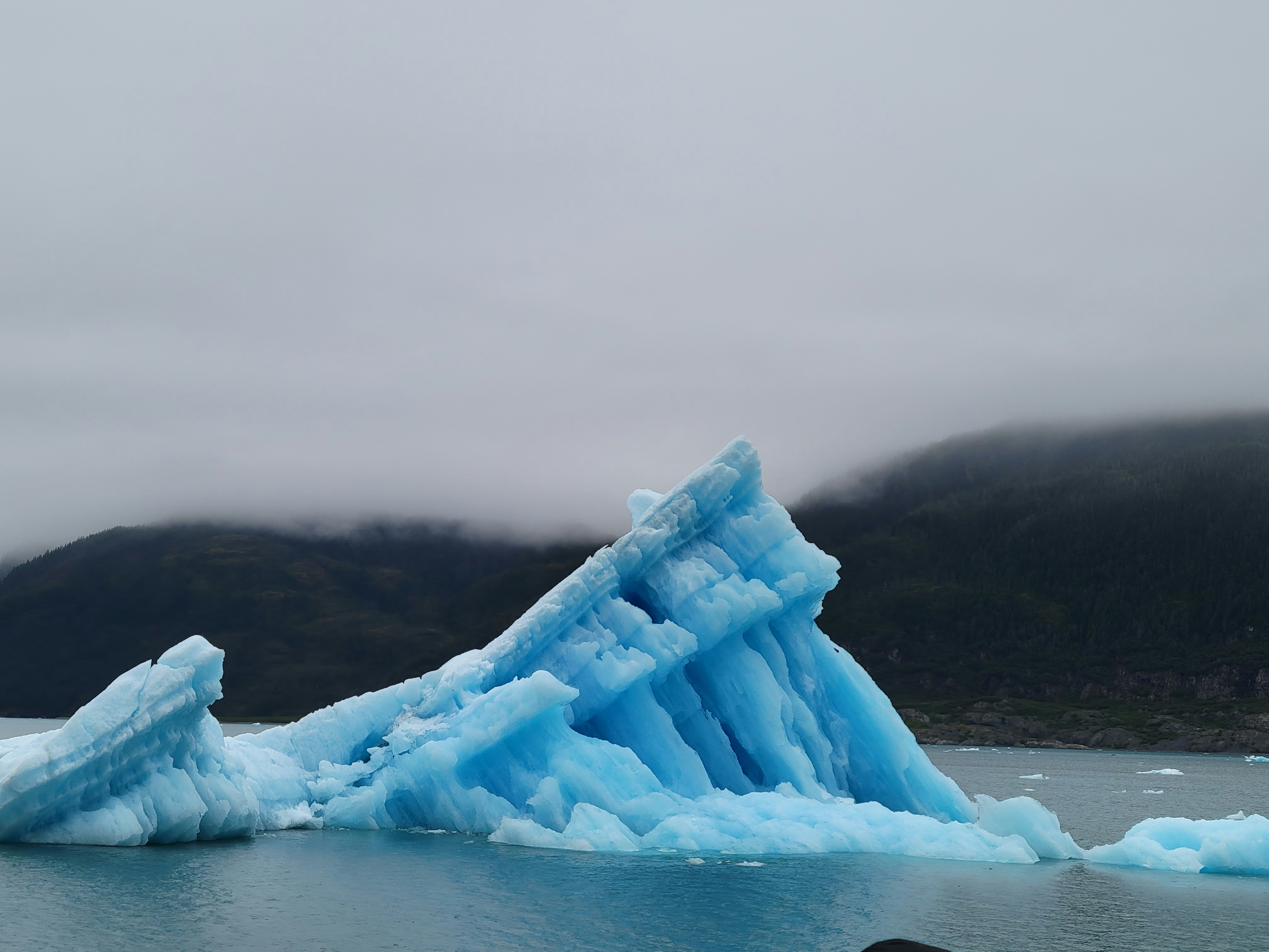 Towering iceberg with striking blue hues emerging from calm waters under a cloudy sky.
