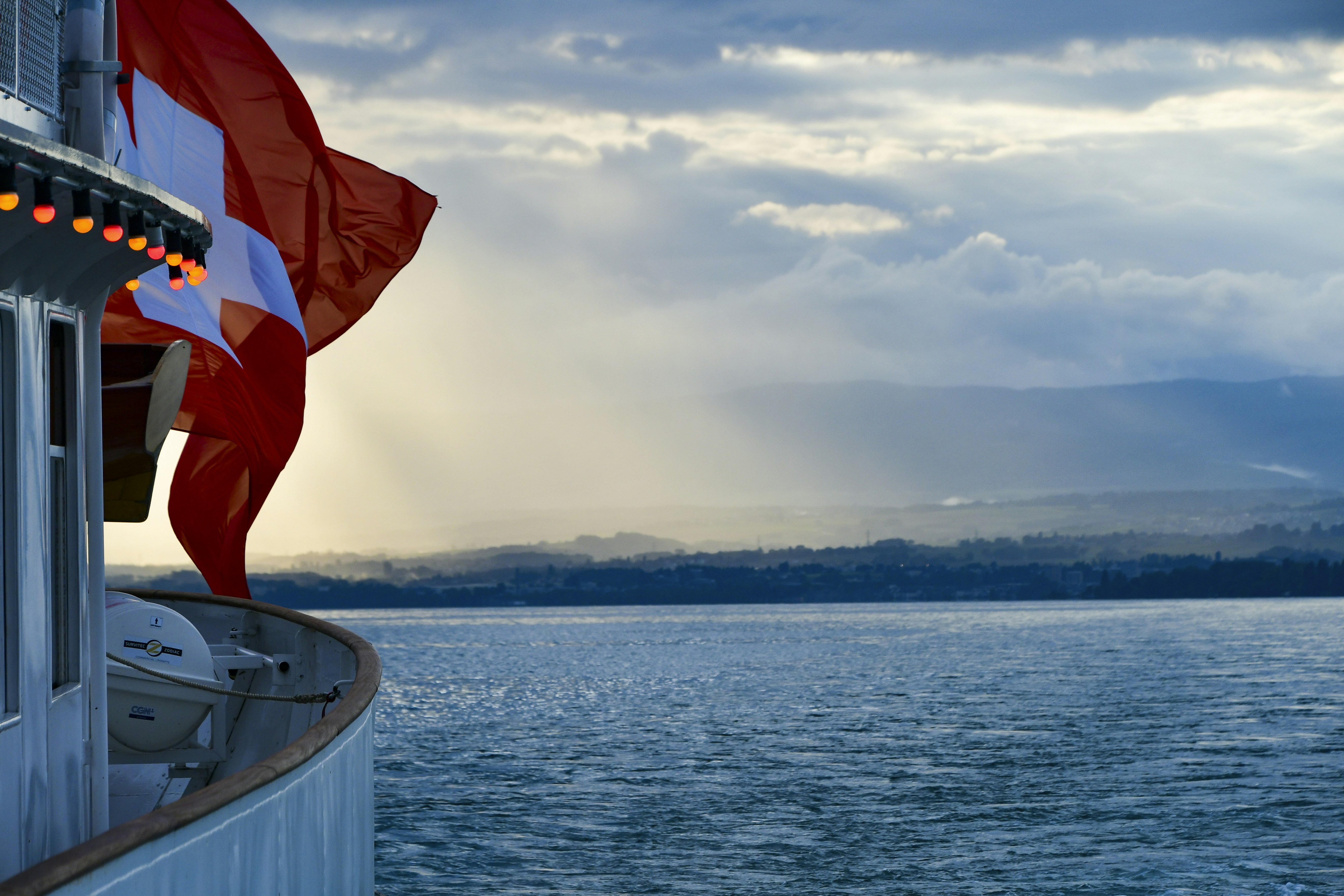 Boat with Swiss flag cruising on a serene lake under a dramatic cloudy sky.