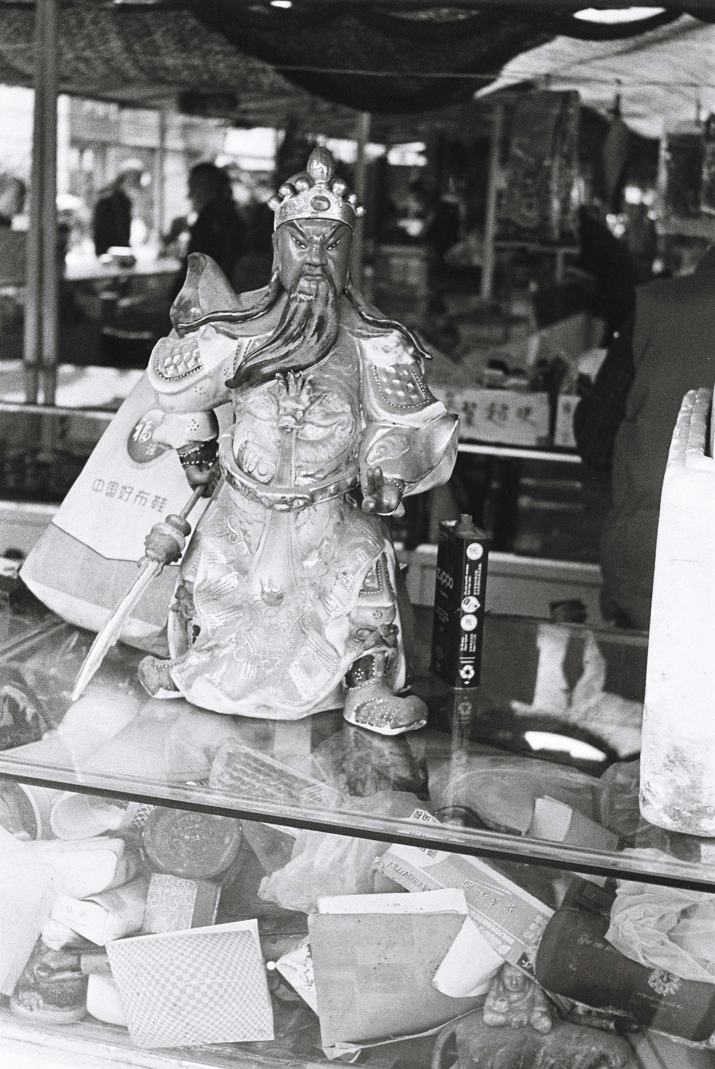 Intricately designed figurine of a warrior, standing prominently on a display table filled with assorted items. The scene captures the vibrant atmosphere of a bustling market.