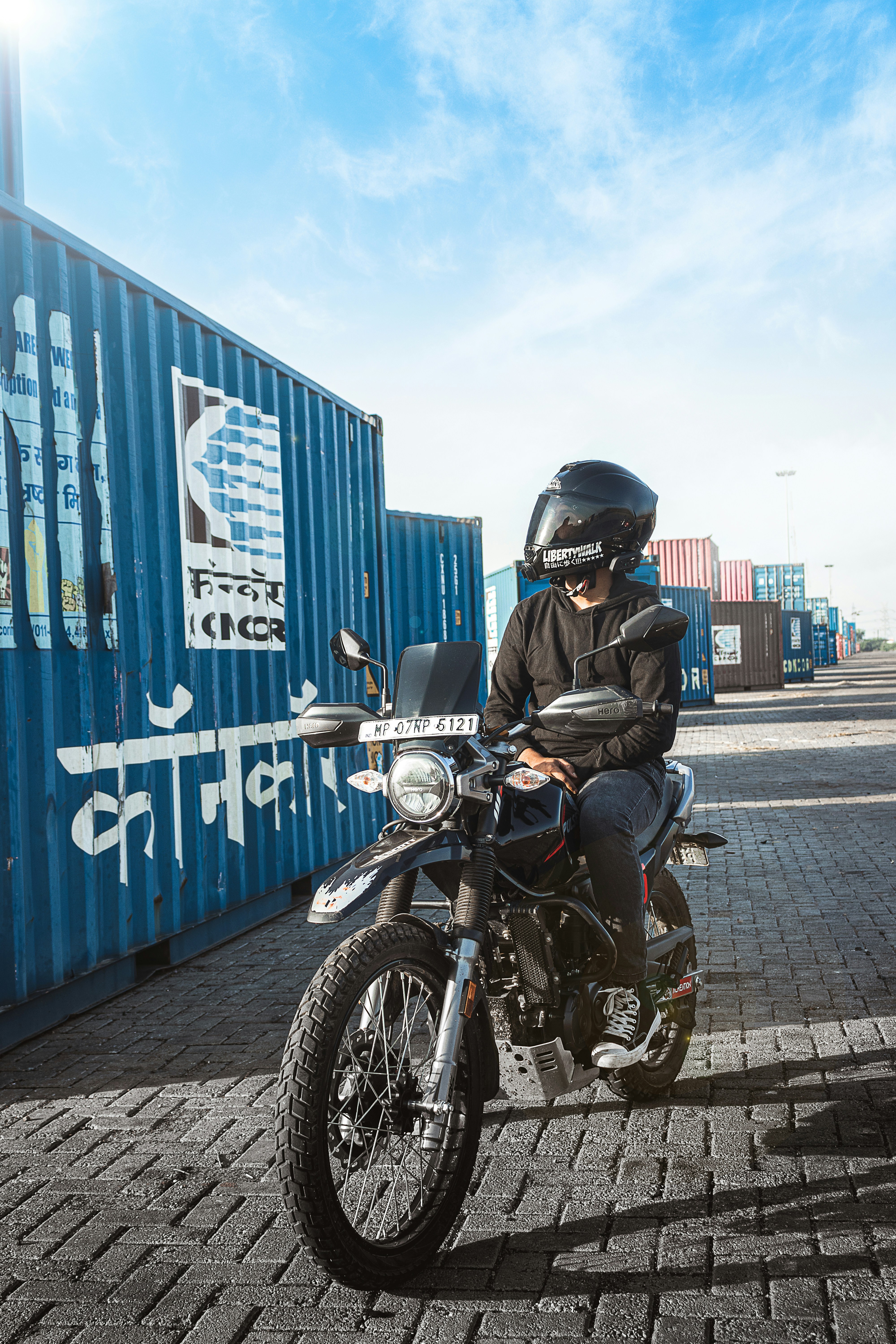 Motorcyclist with helmet pauses beside colorful shipping containers under a bright blue sky.
