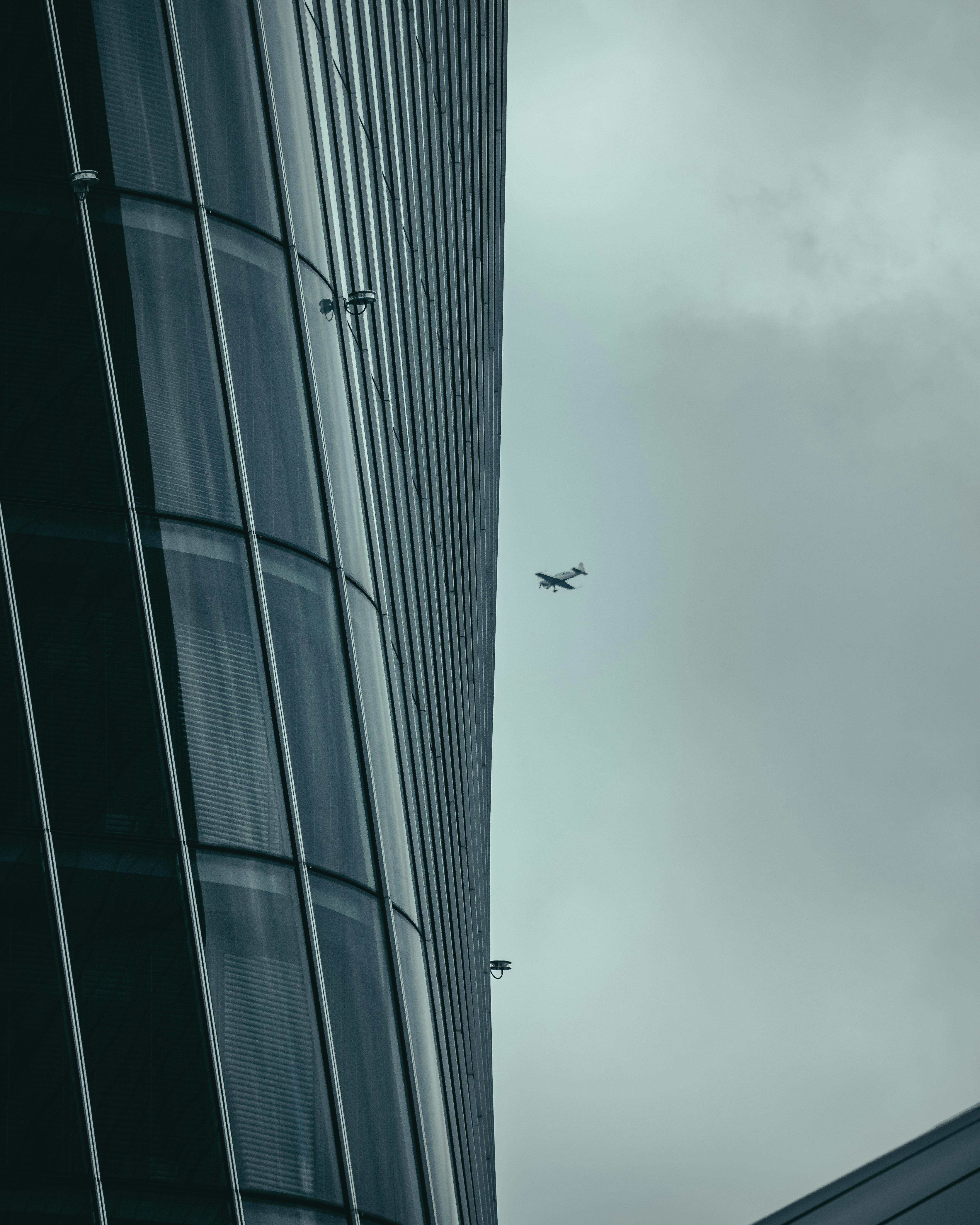 A sleek glass building towers against a moody sky, with a small aircraft flying by, highlighting the intersection of urban design and air travel.