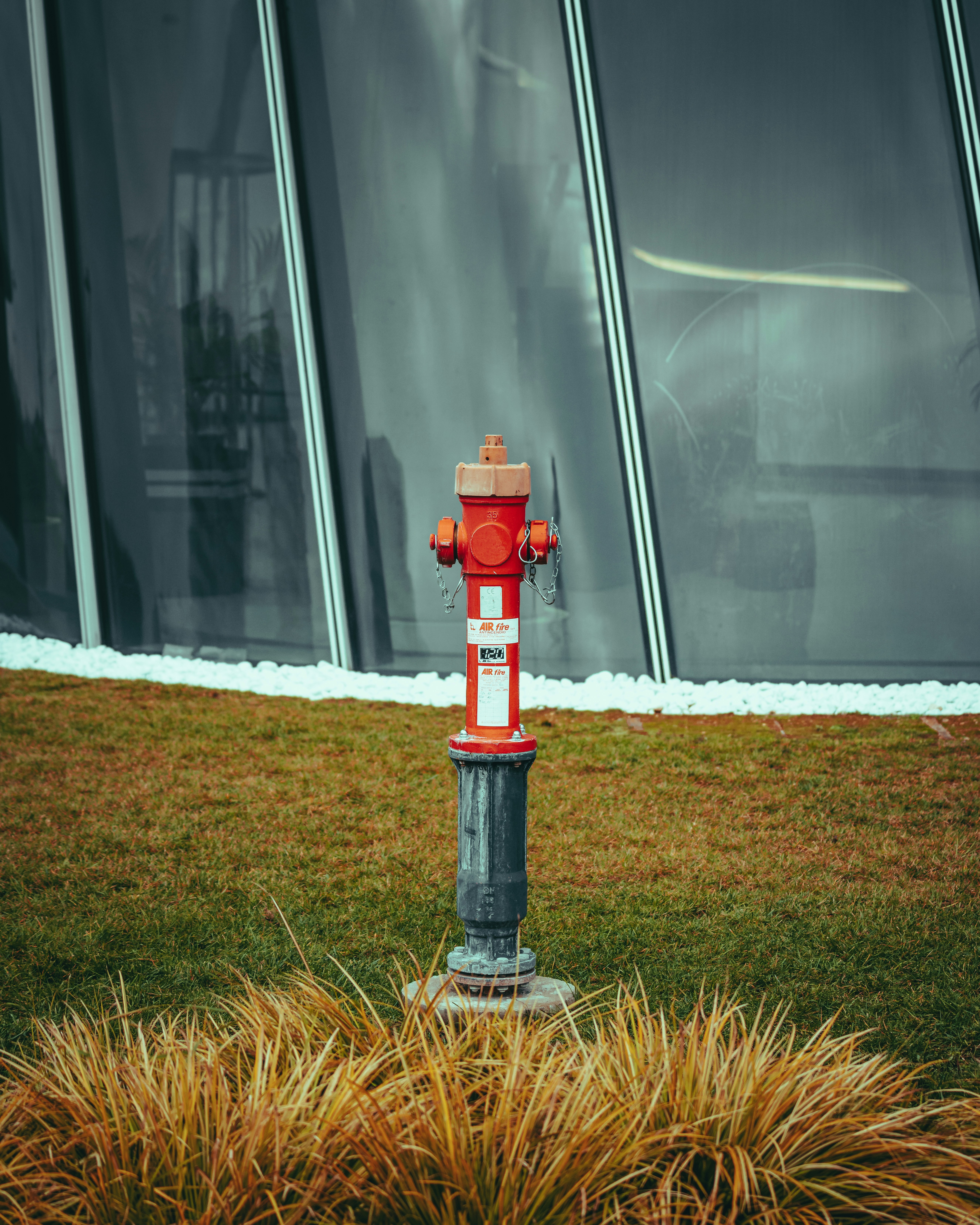 A vibrant red fire hydrant stands prominently on a grassy patch, framed by sleek, modern architecture in the background. Its bright color contrasts sharply with the muted tones of the surroundings.