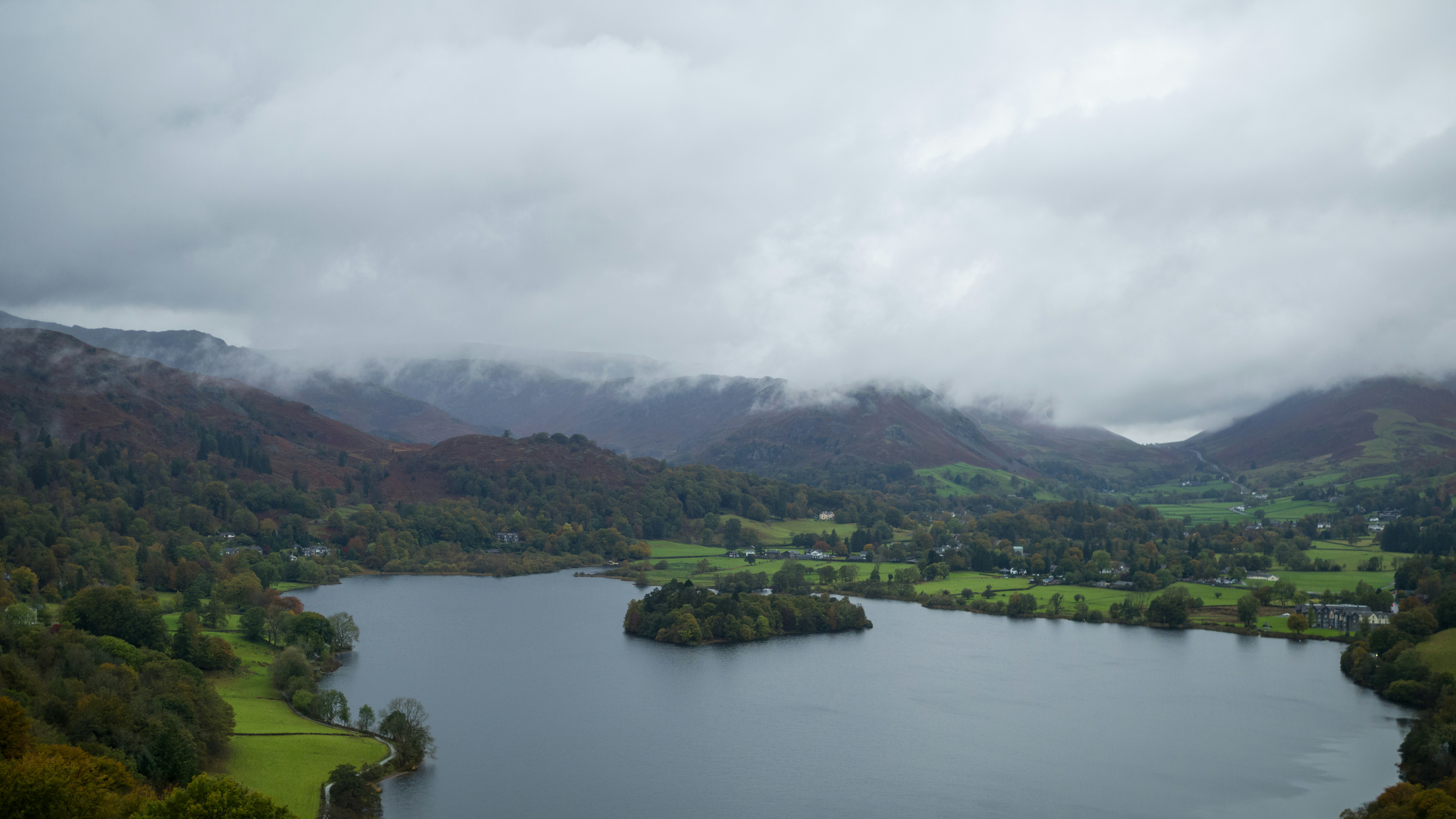 Serene view of a lake surrounded by lush greenery and rolling hills under a cloudy sky. The landscape captures the essence of calmness and natural beauty.