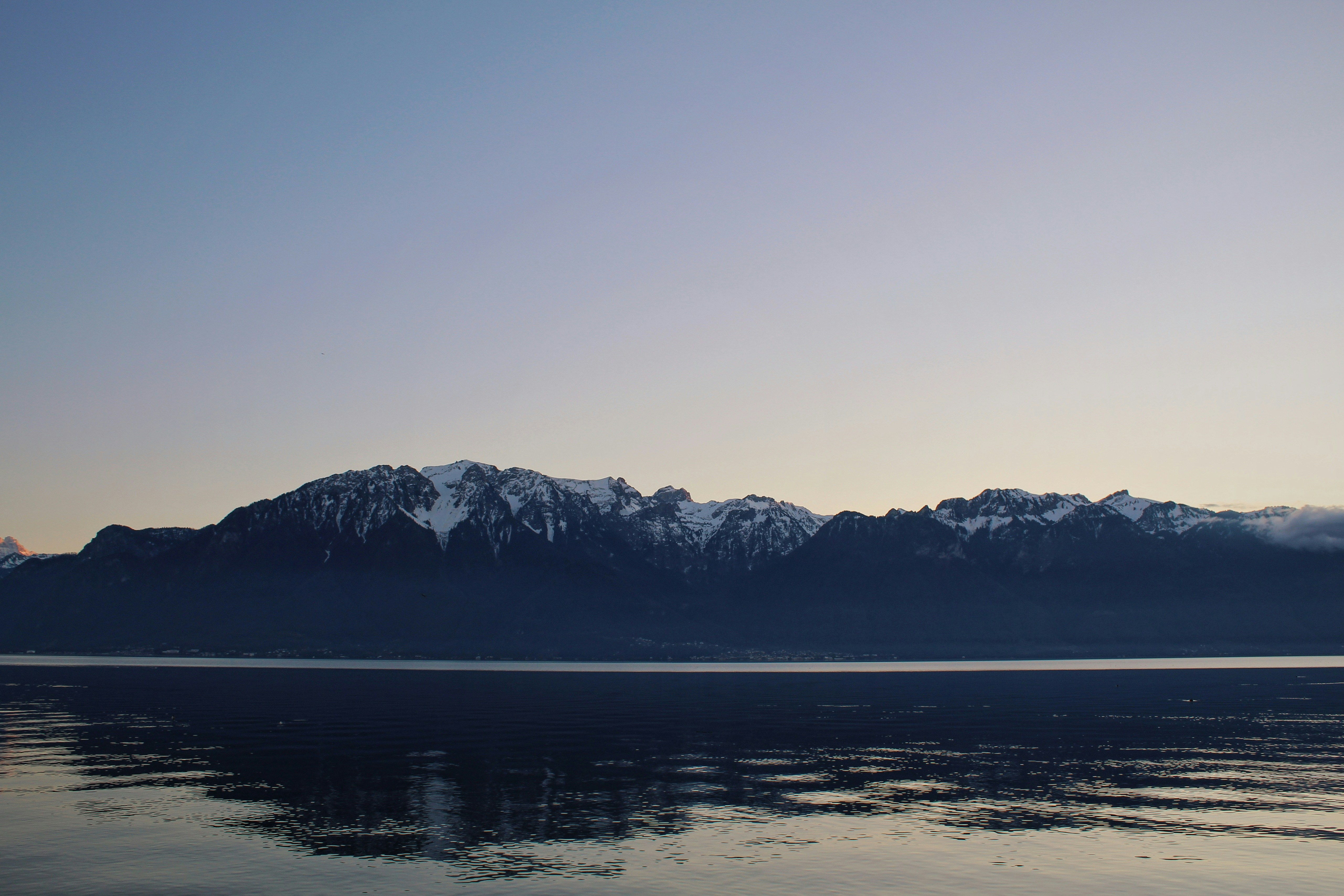 Snow-capped mountains silhouetted against a twilight sky, reflecting on the calm surface of a lake.