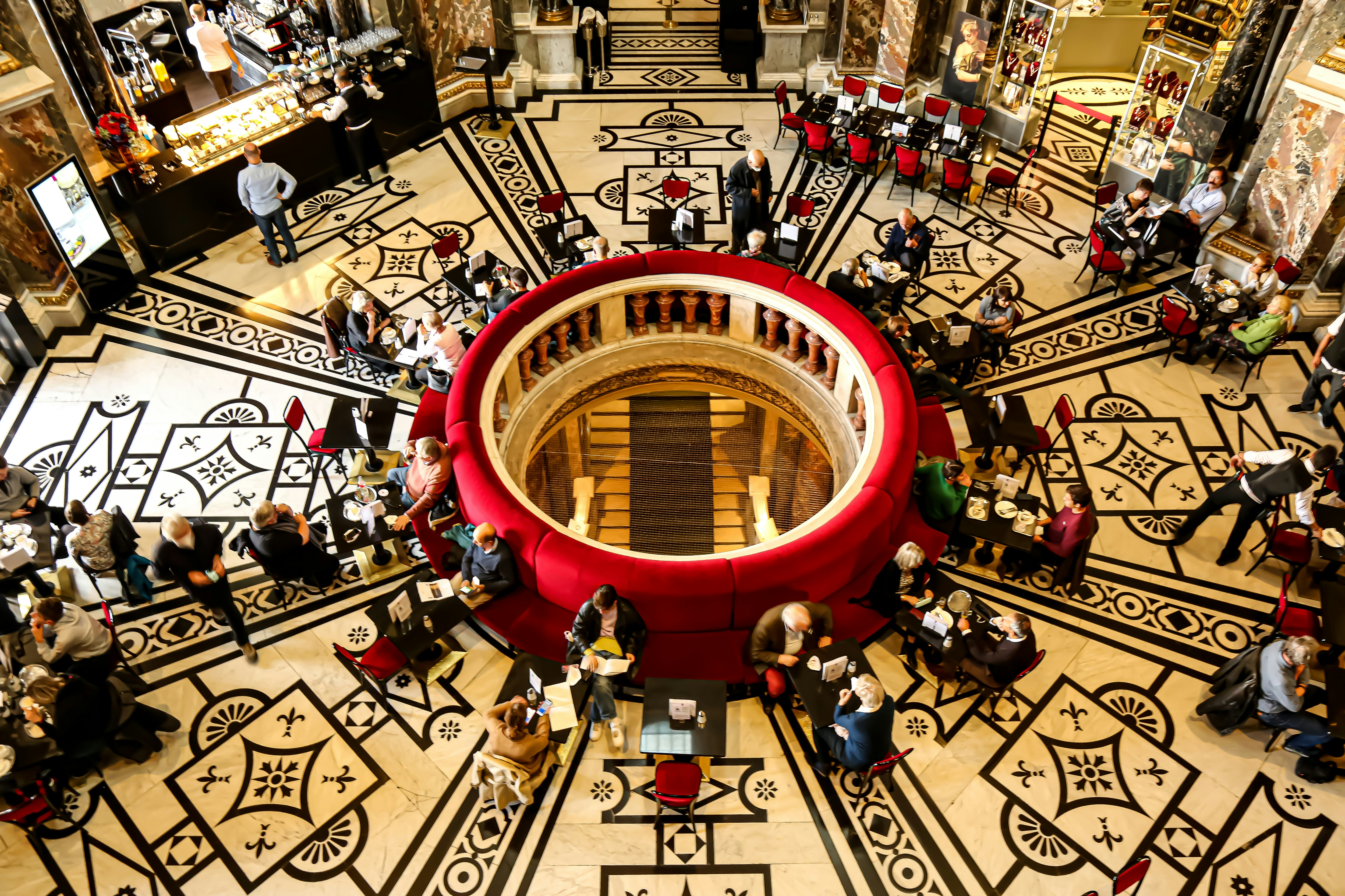 Elegant café scene in a grand atrium, showcasing patrons enjoying their time amidst intricate tile patterns and a striking red circular seating area.