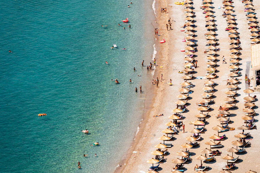 Turquoise sea meets a crowded sandy beach lined with rows of straw umbrellas and sun loungers. Swimmers dot the water near the shoreline.