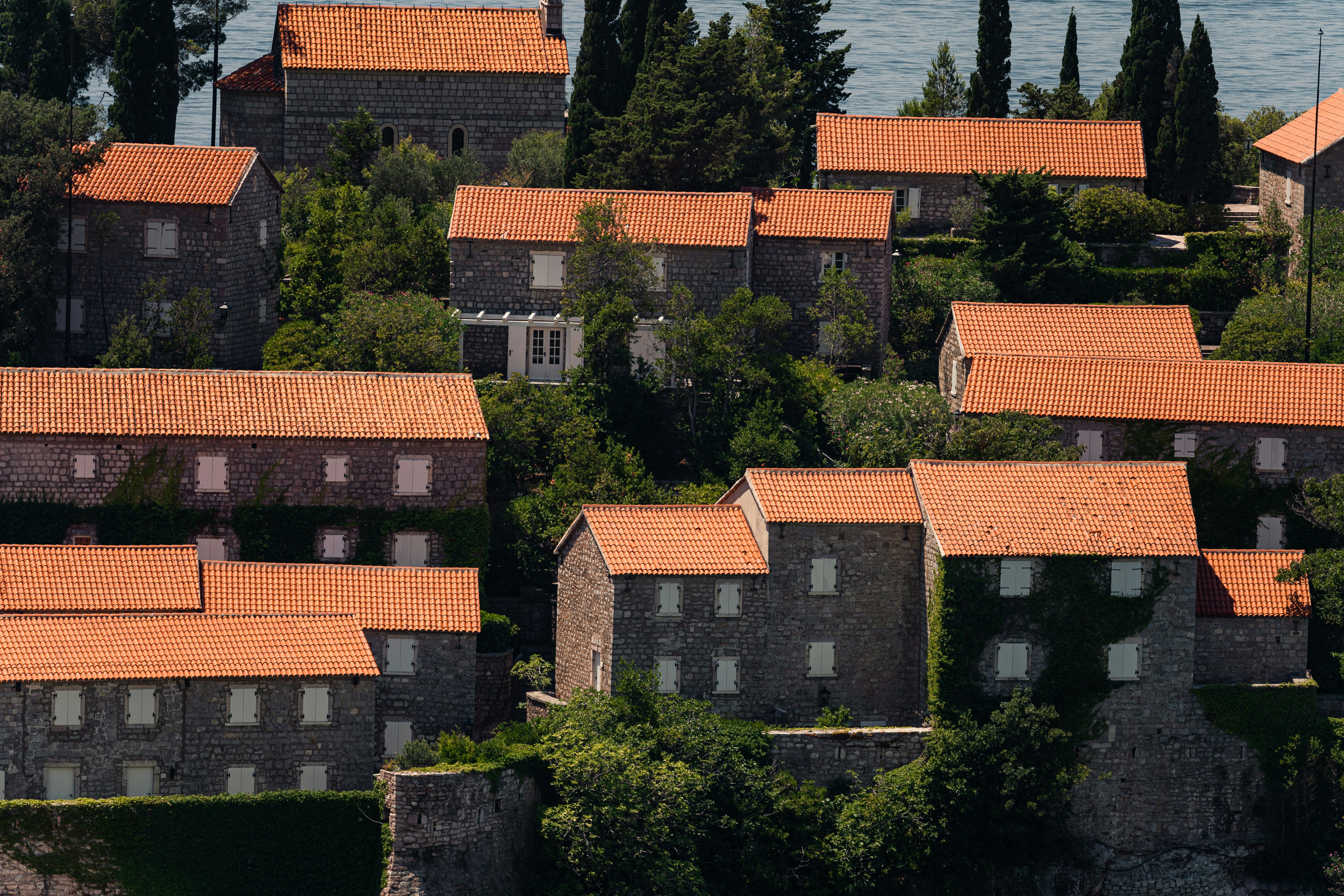 Aerial view of stone houses with terracotta roofs surrounded by lush greenery and a tranquil body of water in the background.