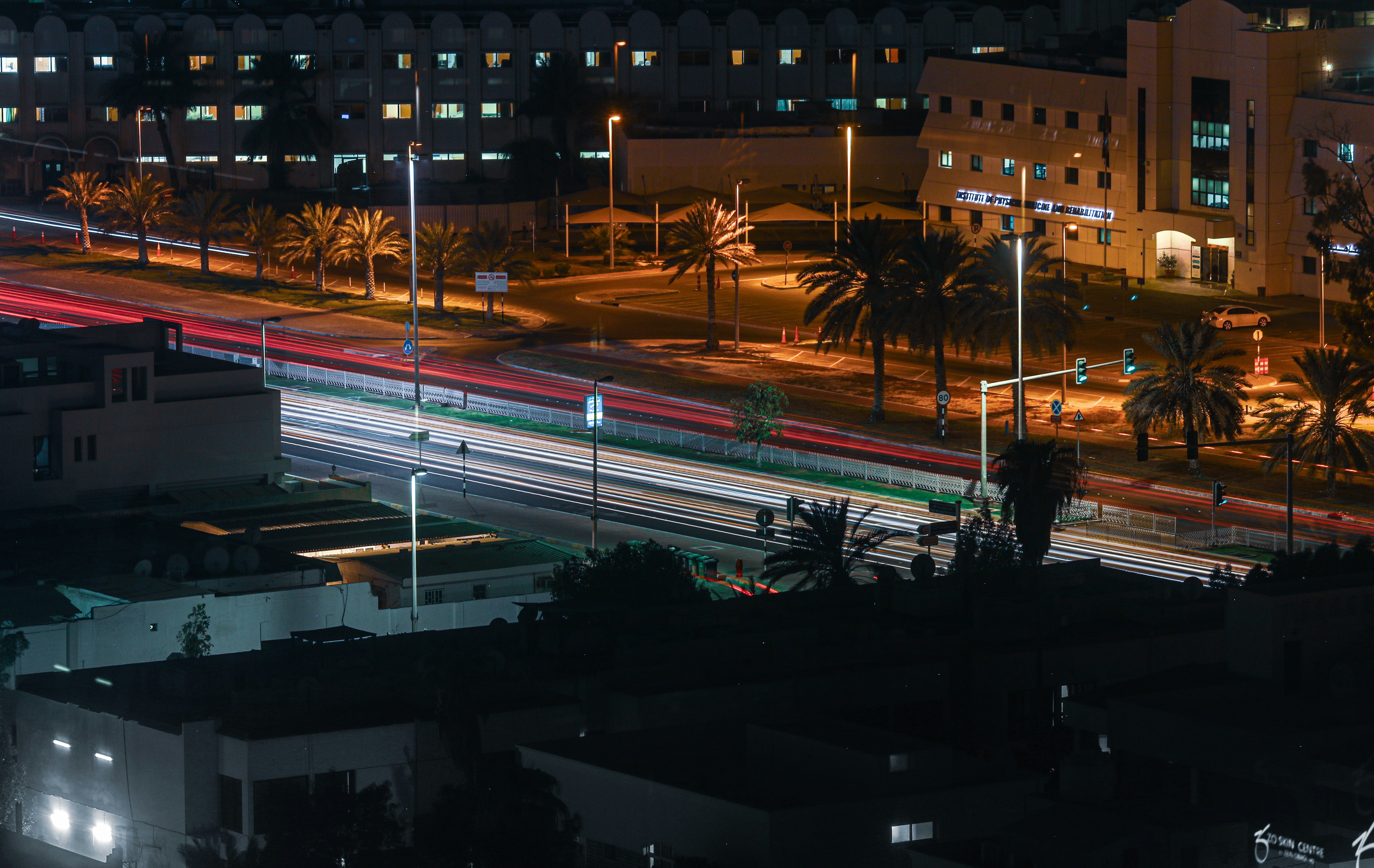 Long exposure capturing light trails of vehicles on a city road, framed by palm trees and urban architecture. The scene showcases the dynamic energy of nighttime traffic.