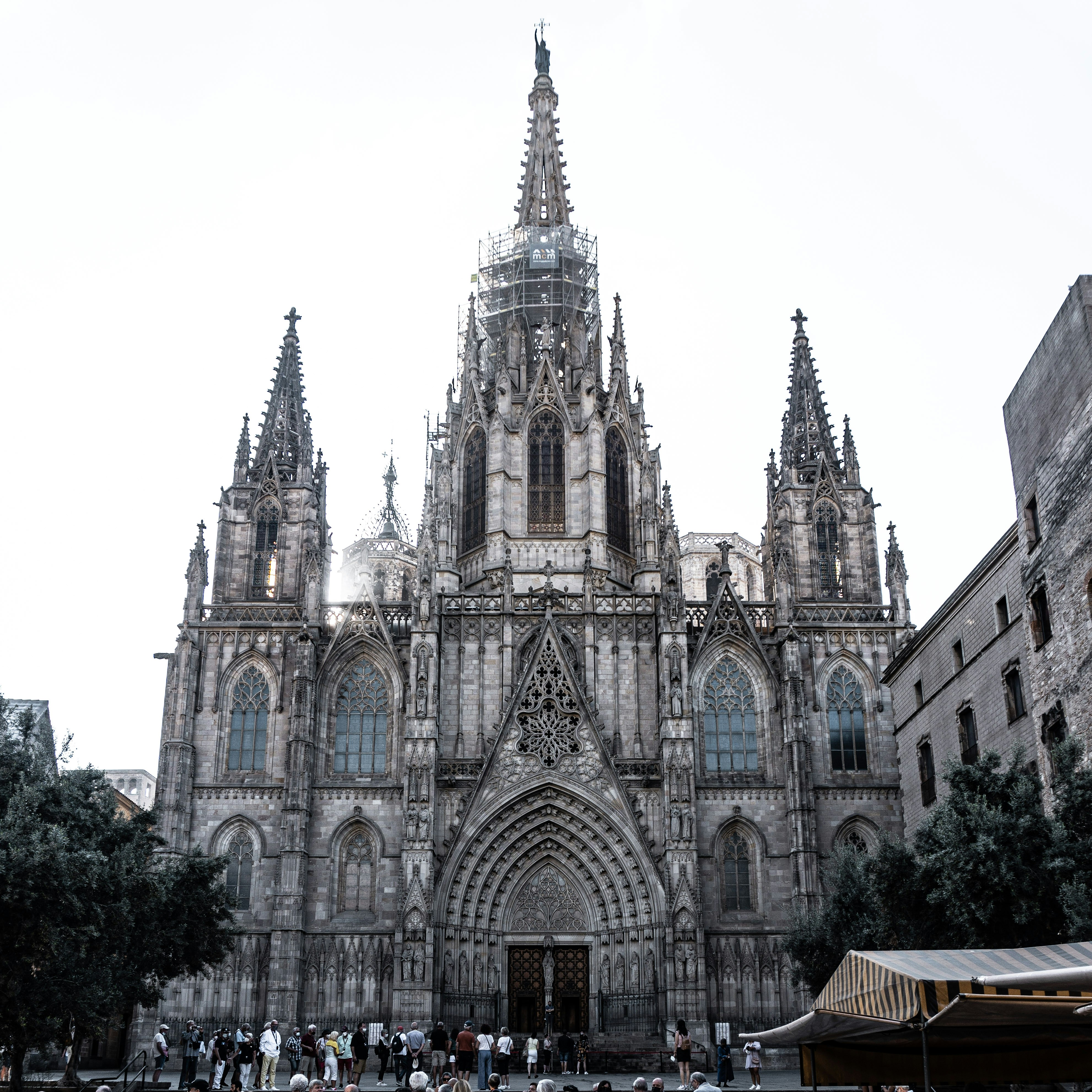 Imposing Gothic cathedral with intricate spires and detailed facade, surrounded by visitors in a bustling square. Scaffolding hints at ongoing restoration efforts.