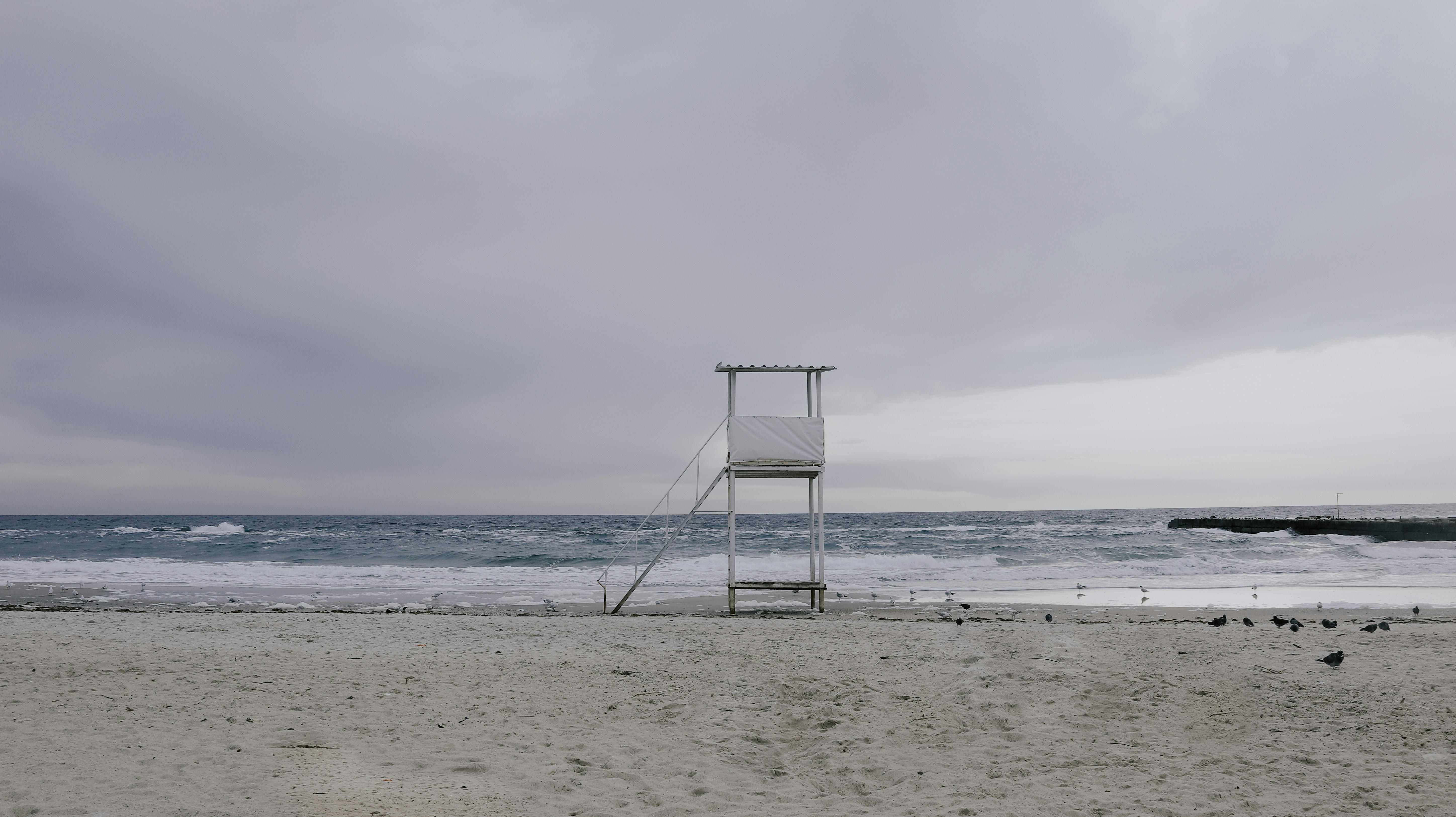 A lifeguard tower stands alone on a sandy beach, overlooking the turbulent sea under a cloudy sky.