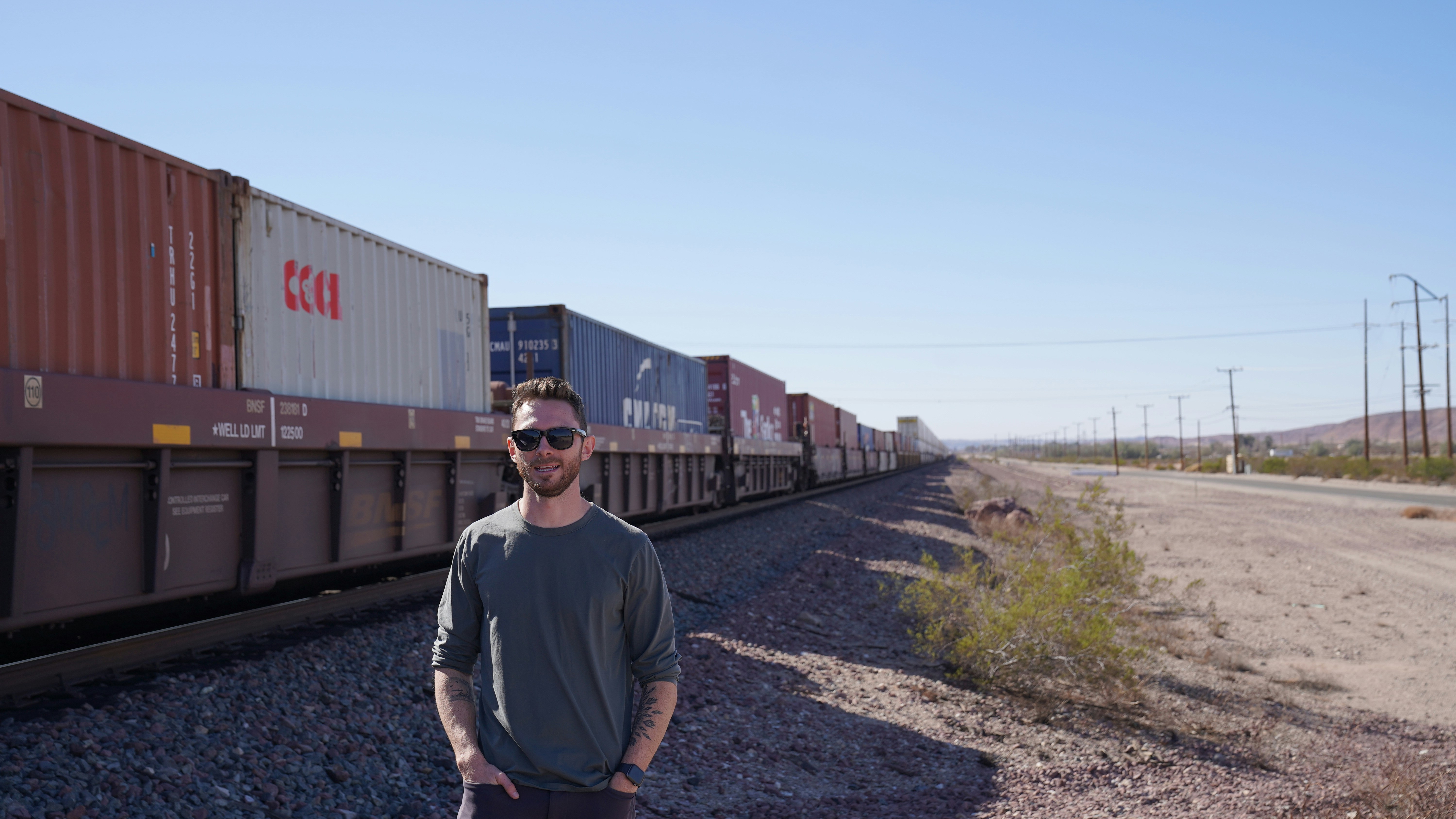 A man stands confidently beside a line of freight containers, showcasing the intersection of human presence and industrial transport.