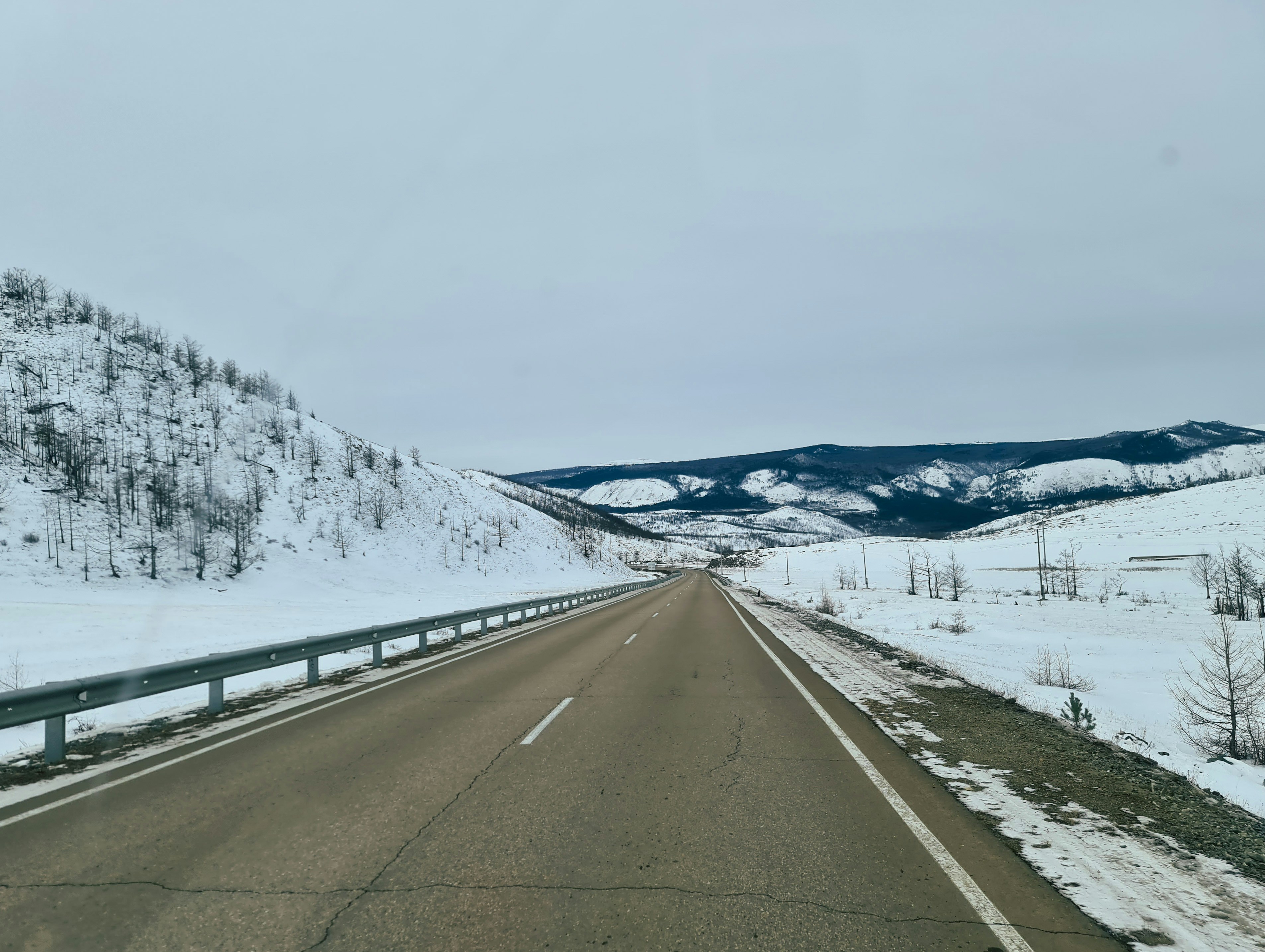 Long, empty highway stretching through snow-covered hills under an overcast sky.