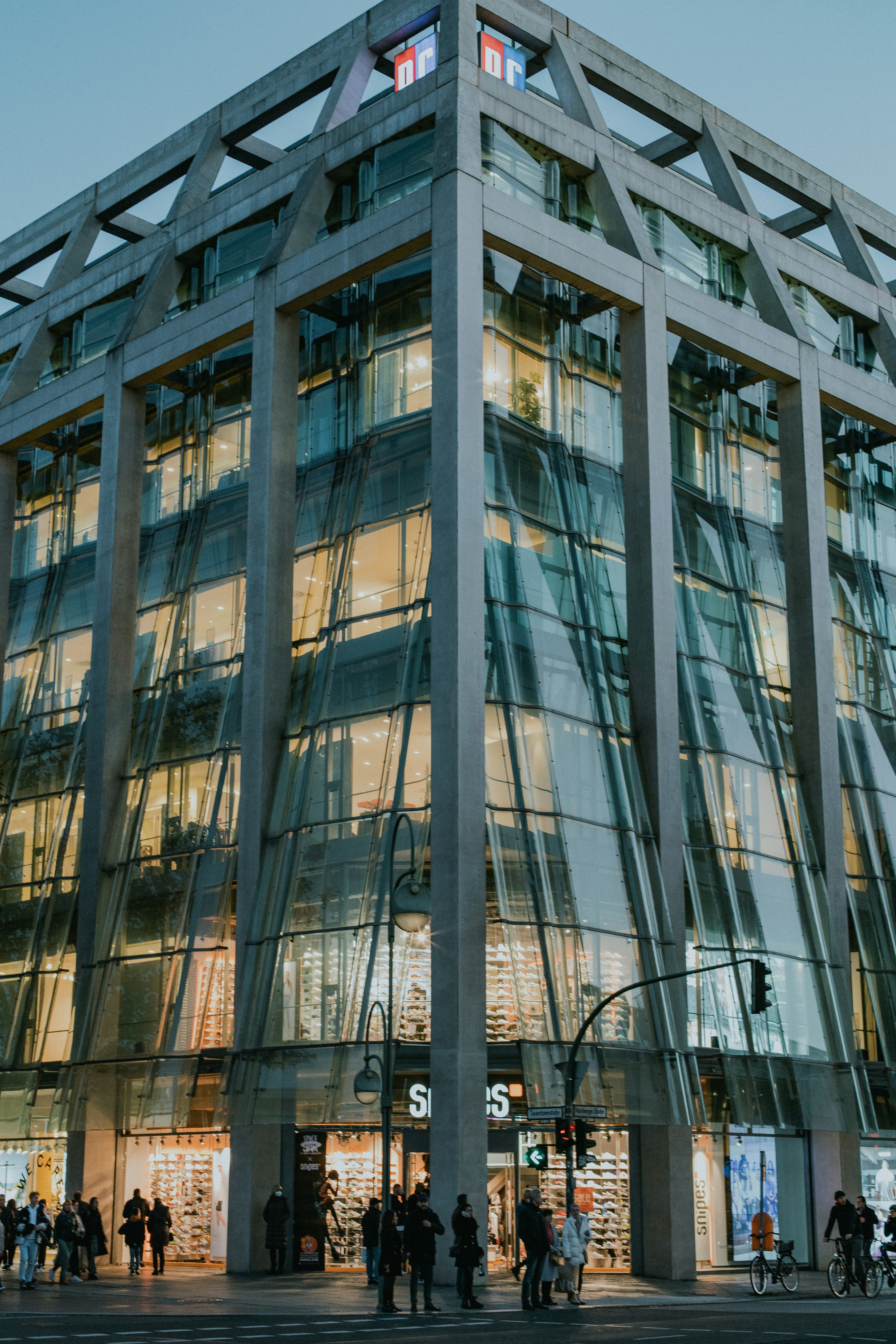 Contemporary building featuring a striking glass facade with intricate reflections, illuminated by warm interior lights. A bustling street scene unfolds at its base.