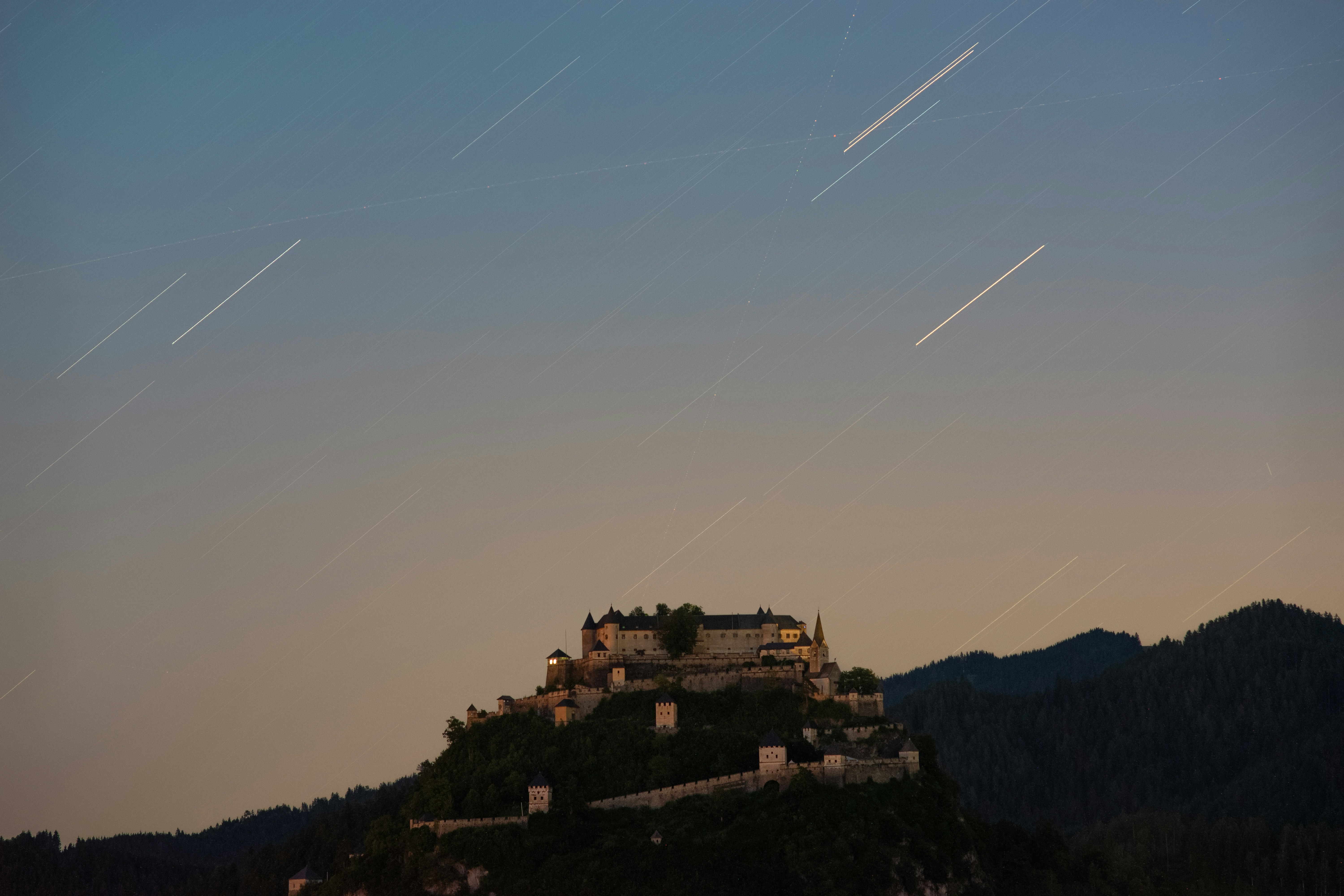 Star trails illuminate a historic fortress perched atop a hill, set against a twilight sky. The image captures the serene blend of nature and architecture.
