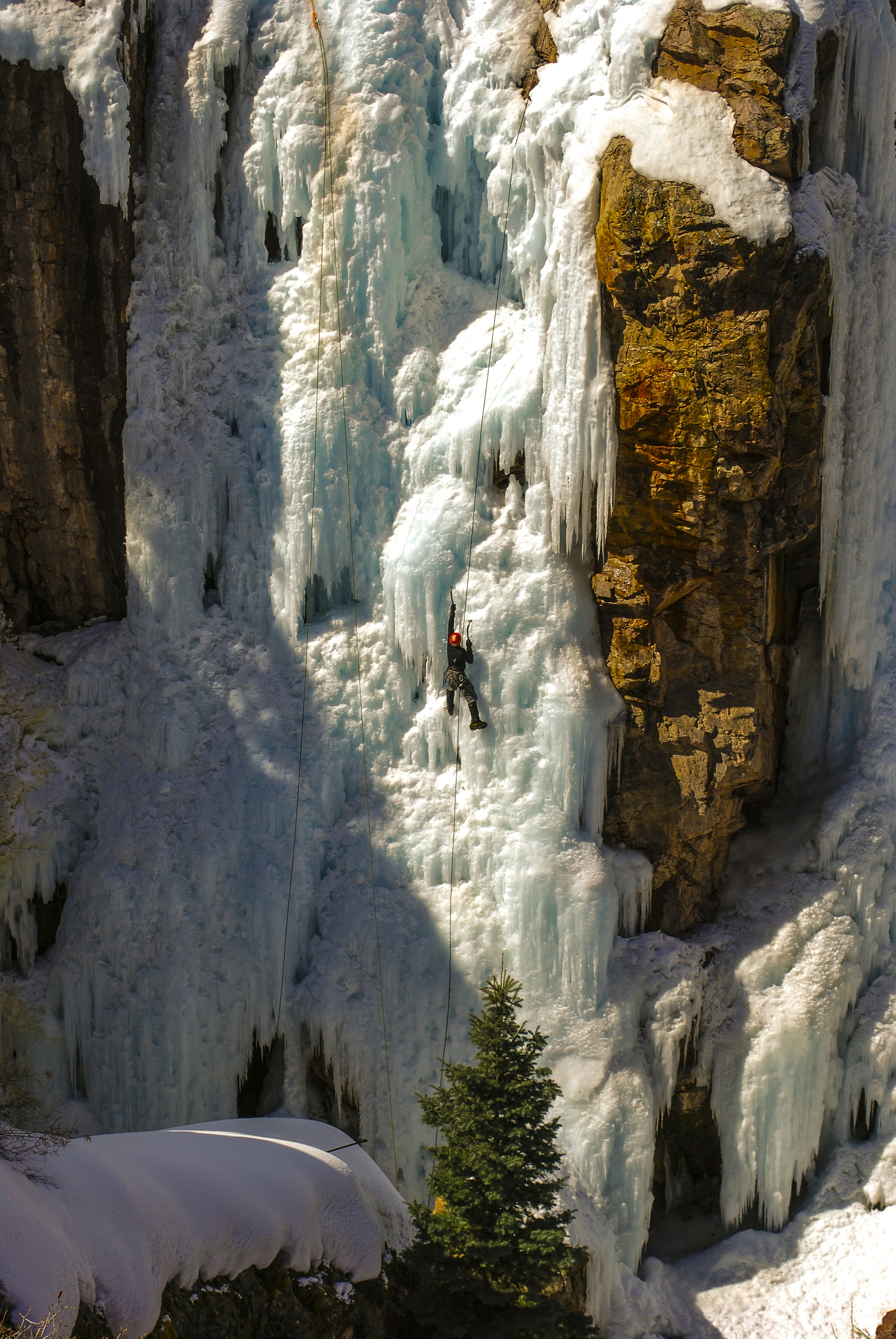 Ice climber scaling a frozen waterfall surrounded by rugged cliffs and snow-covered terrain.