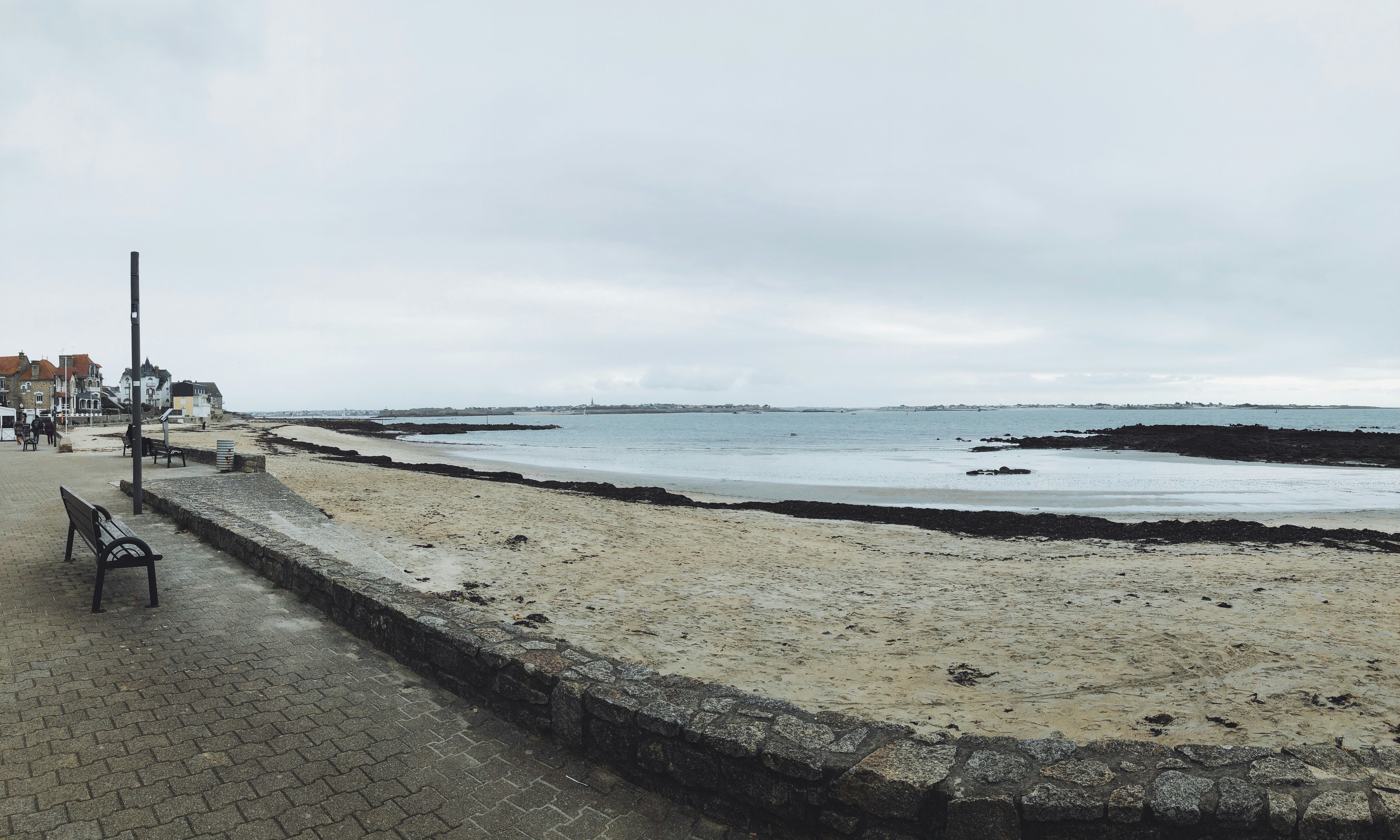 Panoramic view of a tranquil beach with gentle waves and a sandy shore, framed by a stone walkway and distant coastal buildings.