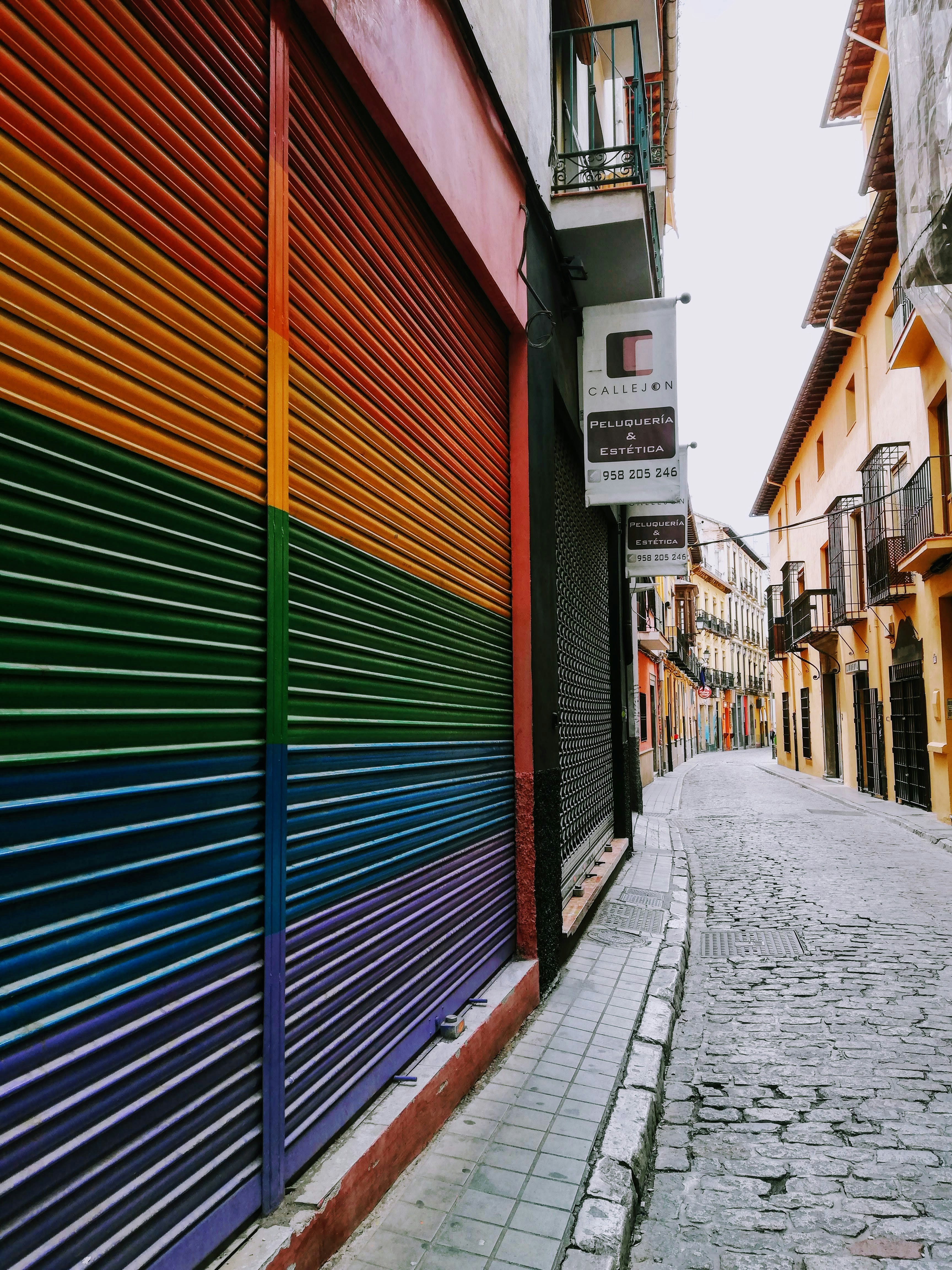 Rainbow-striped shutter on the left frames a sunlit cobblestone street that recedes between warm buildings.