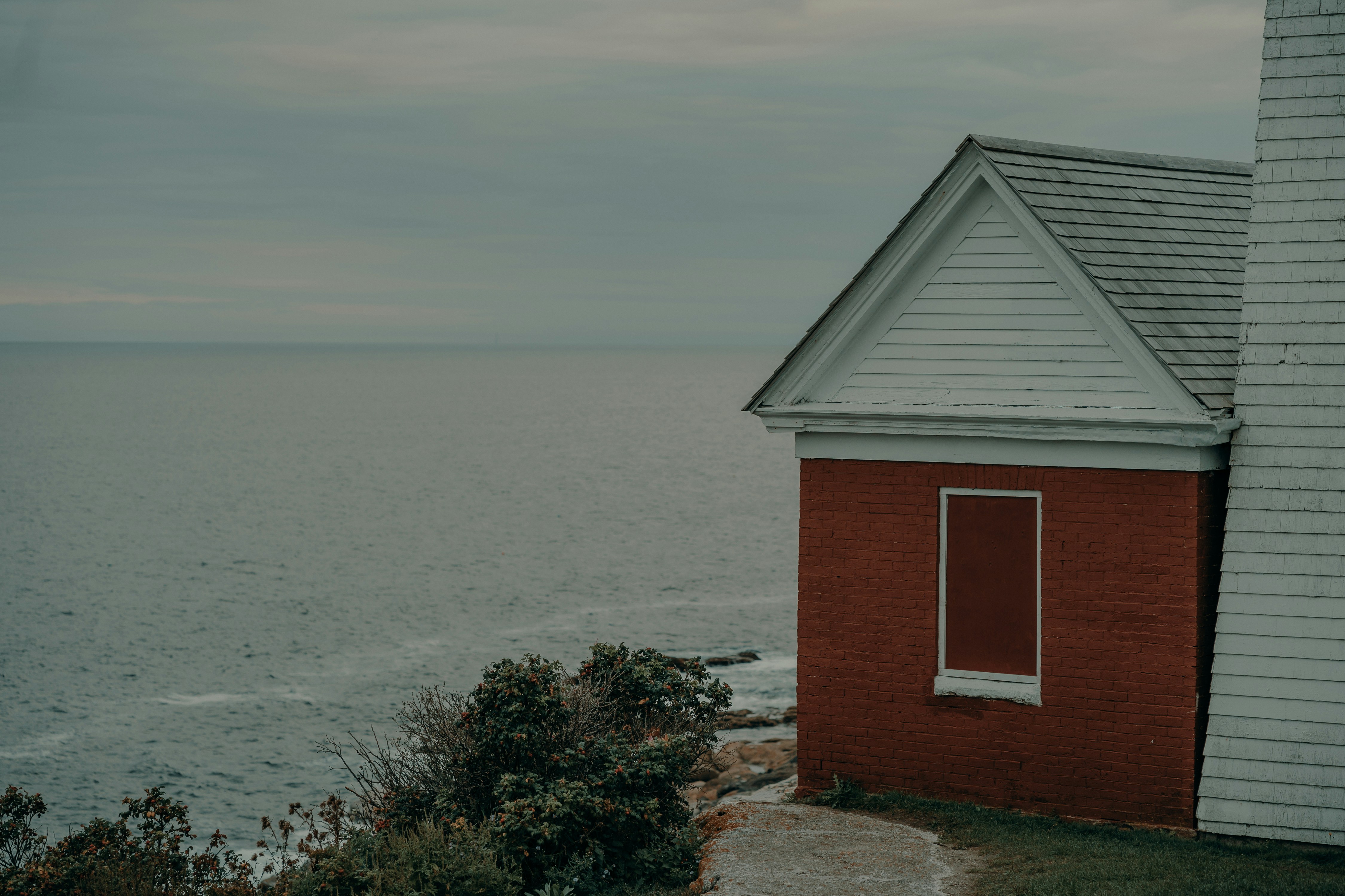 Coastal red cottage stands against a moody seascape, with a hint of nature framing its base. The structure's weathered charm evokes a sense of solitude.