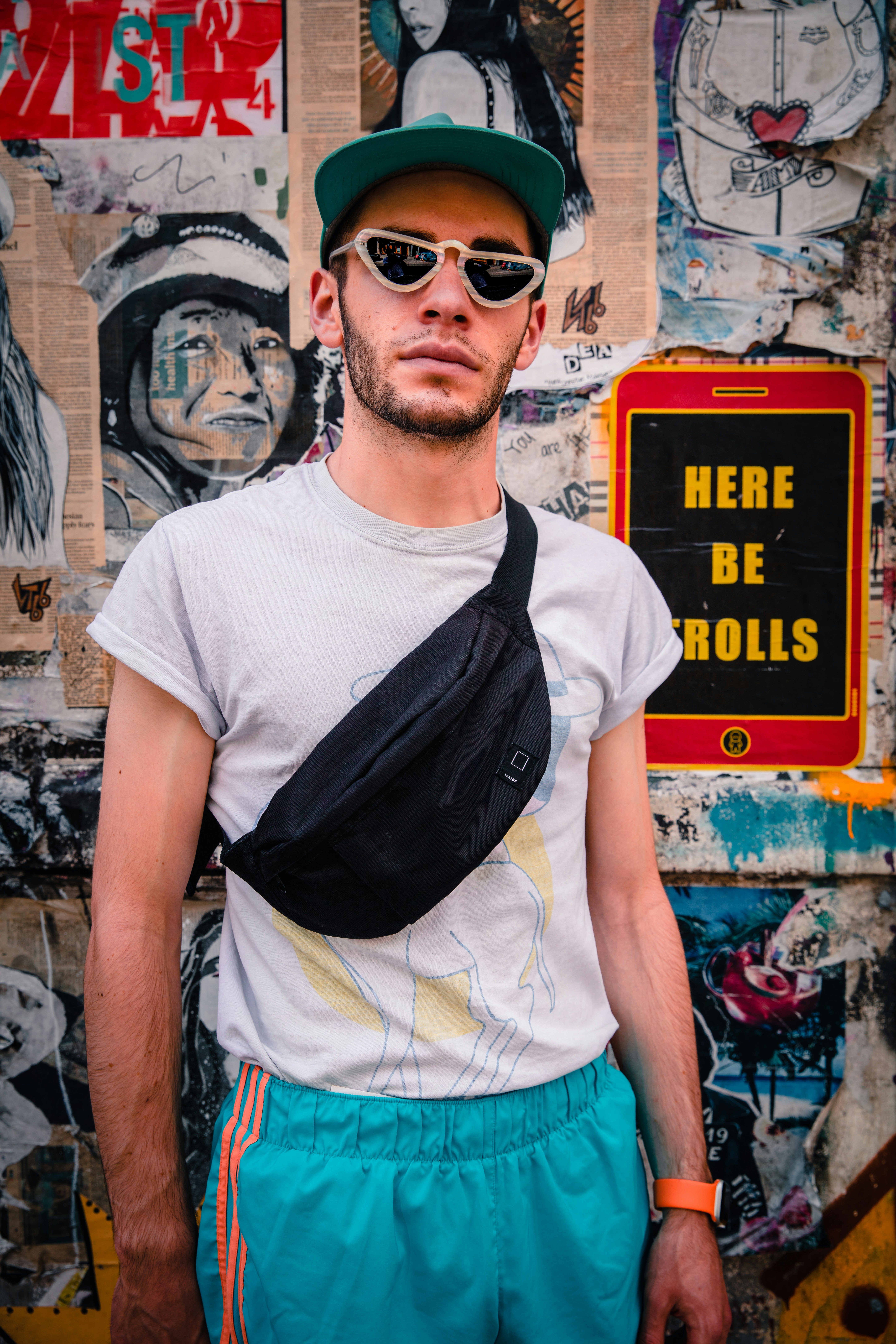 Man in sunglasses and a fanny pack stands in front of a vibrant, graffiti-covered wall featuring various artworks and a bold sign. 