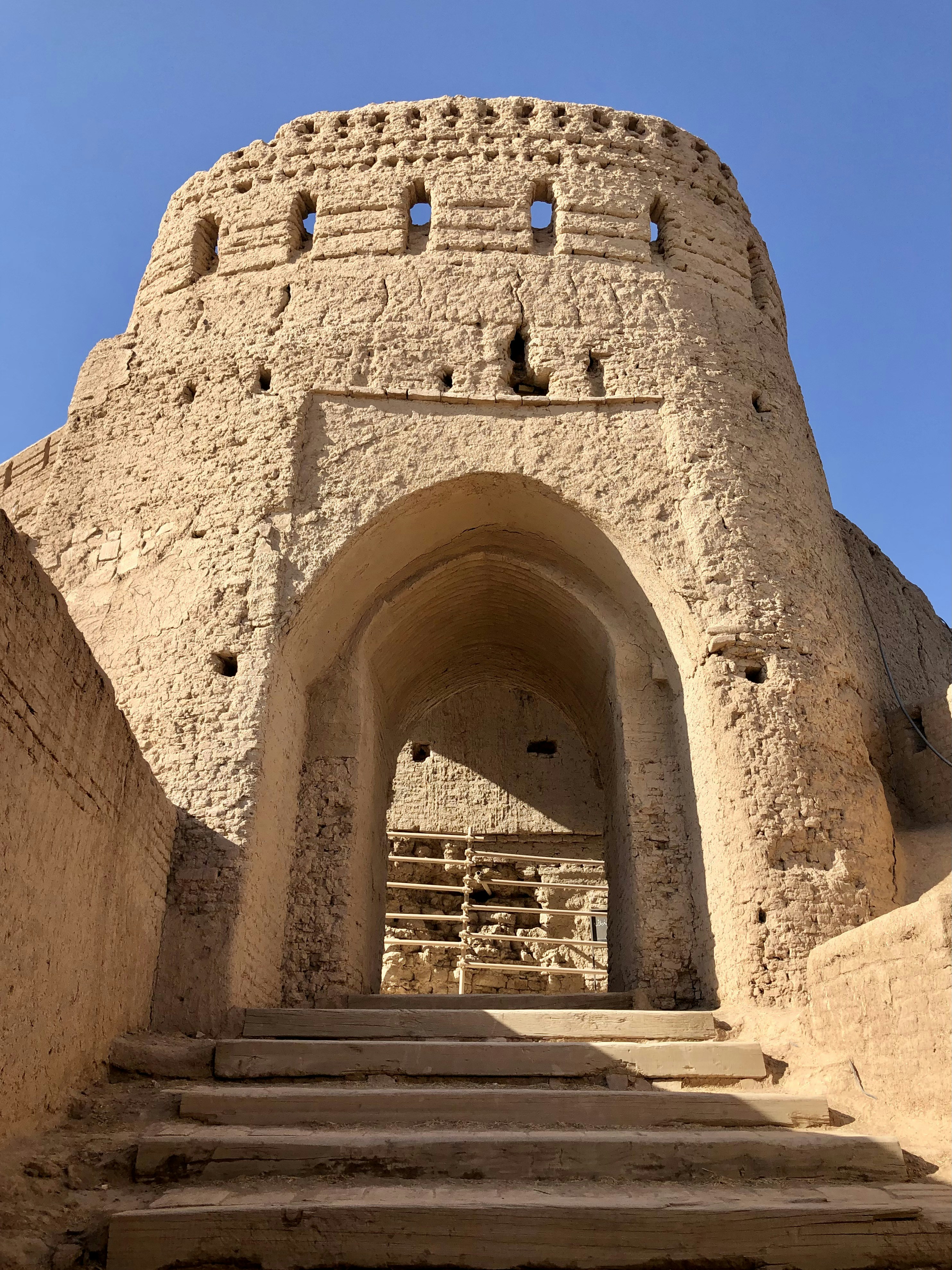 Weathered stone tower with arched entrance and small windows, surrounded by earthen walls. Steps lead up to the entrance, showcasing ancient craftsmanship.