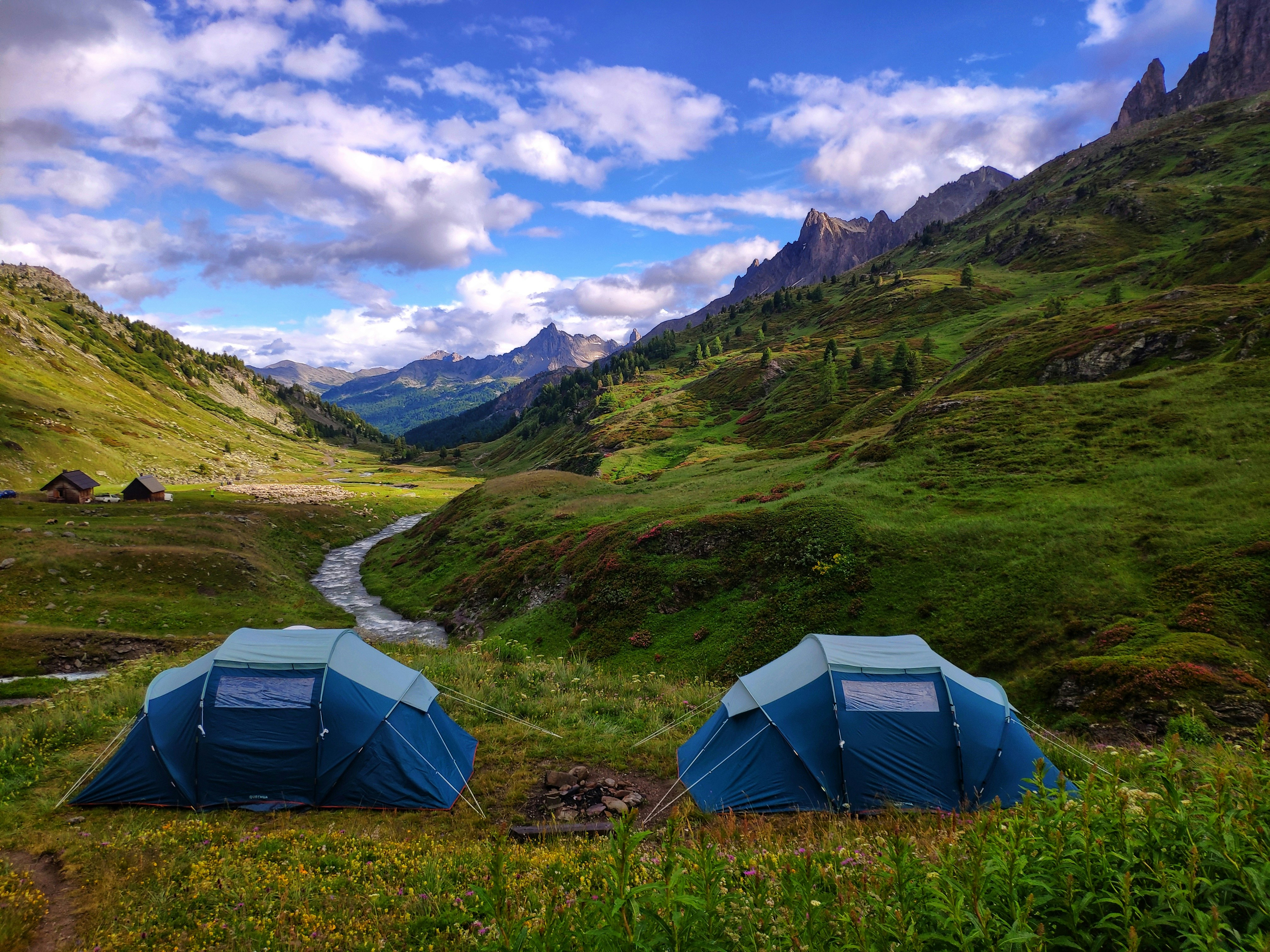Two blue tents sit pitched in a vibrant alpine meadow with a meandering stream, framed by rugged mountains under a bright, partly cloudy sky.