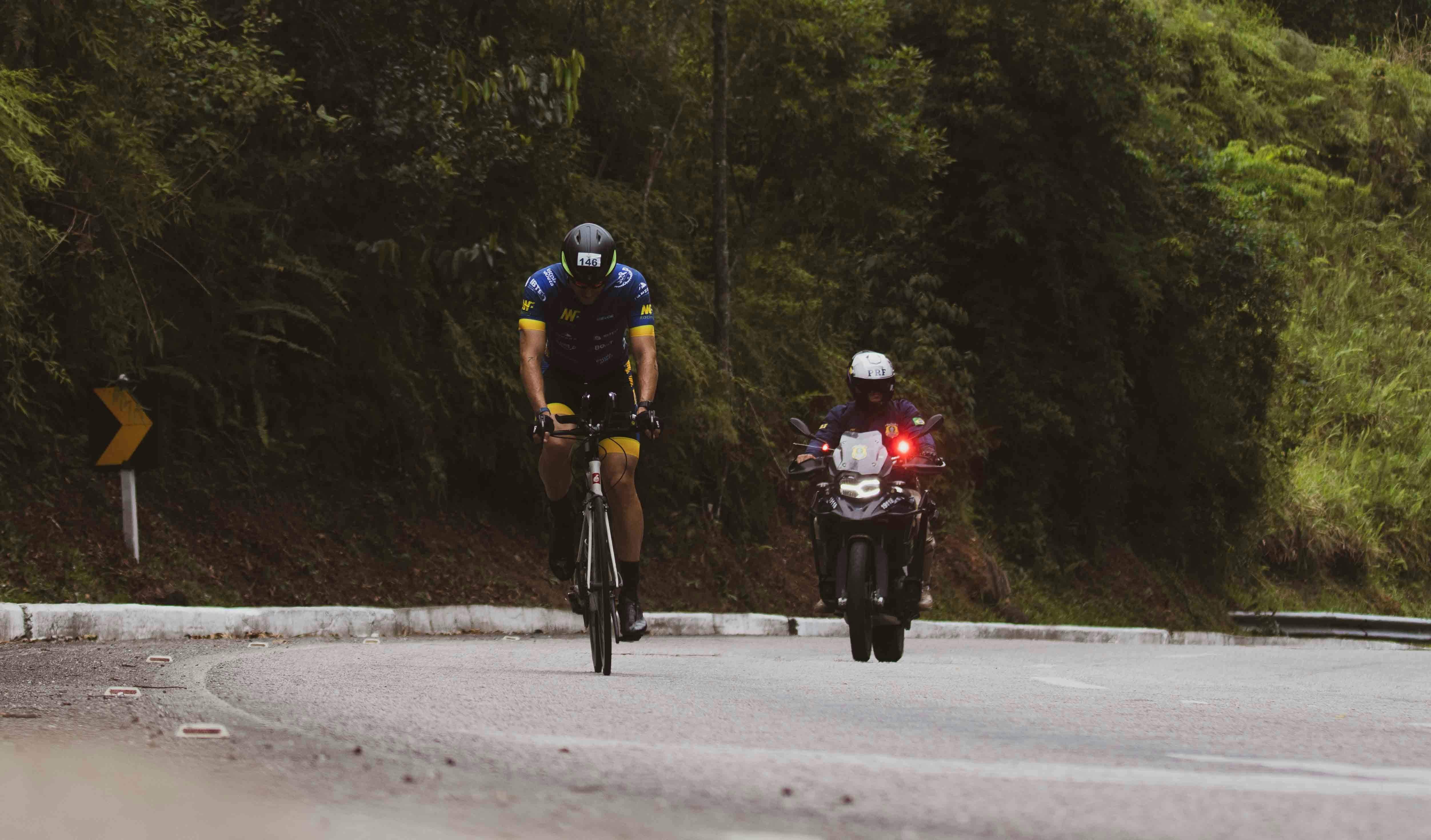 Cyclist and motorcyclist riding side by side on a curving road surrounded by lush greenery.
