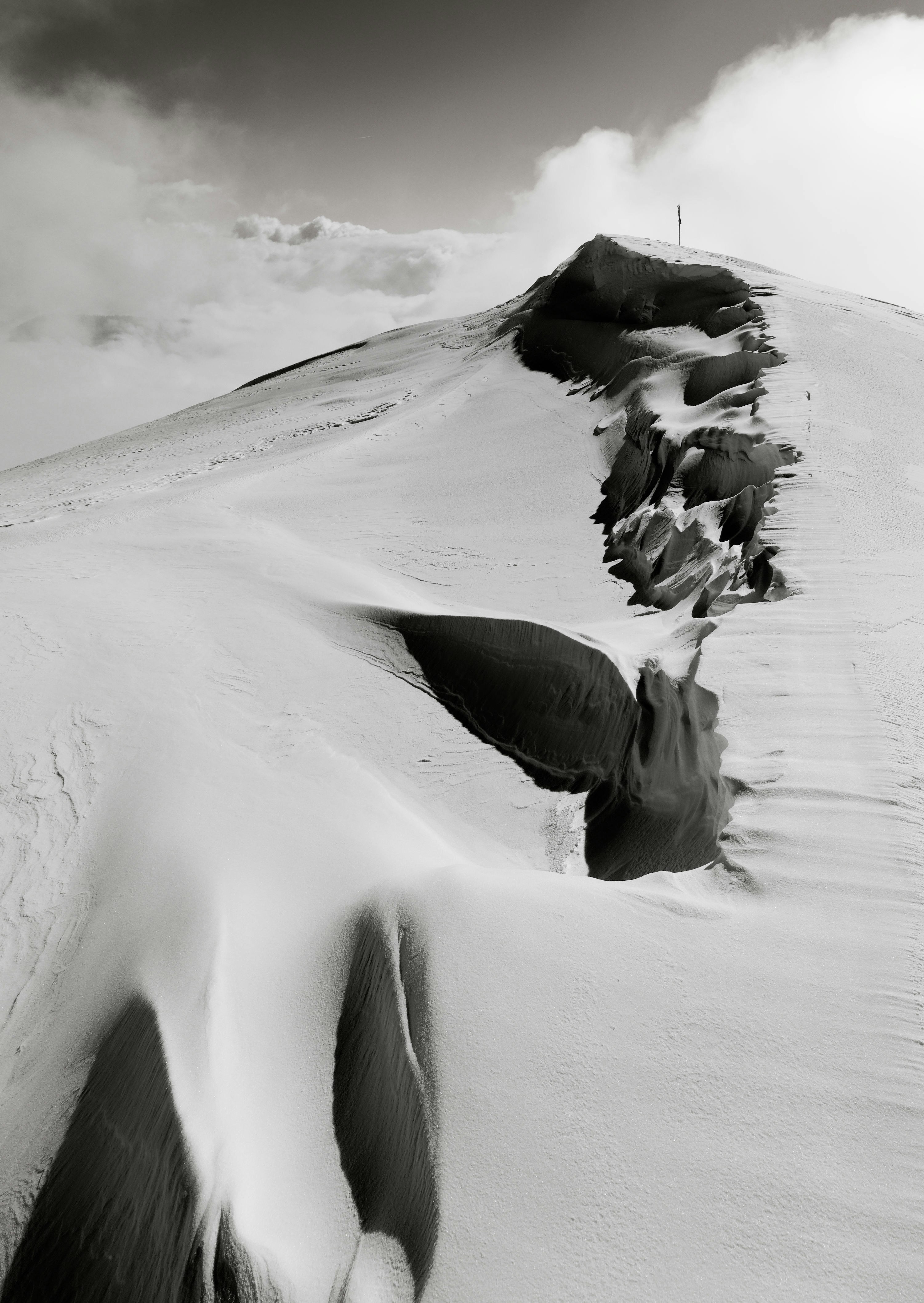 Snow-covered mountain ridge with dramatic crevices and flowing snow patterns under a cloudy sky.